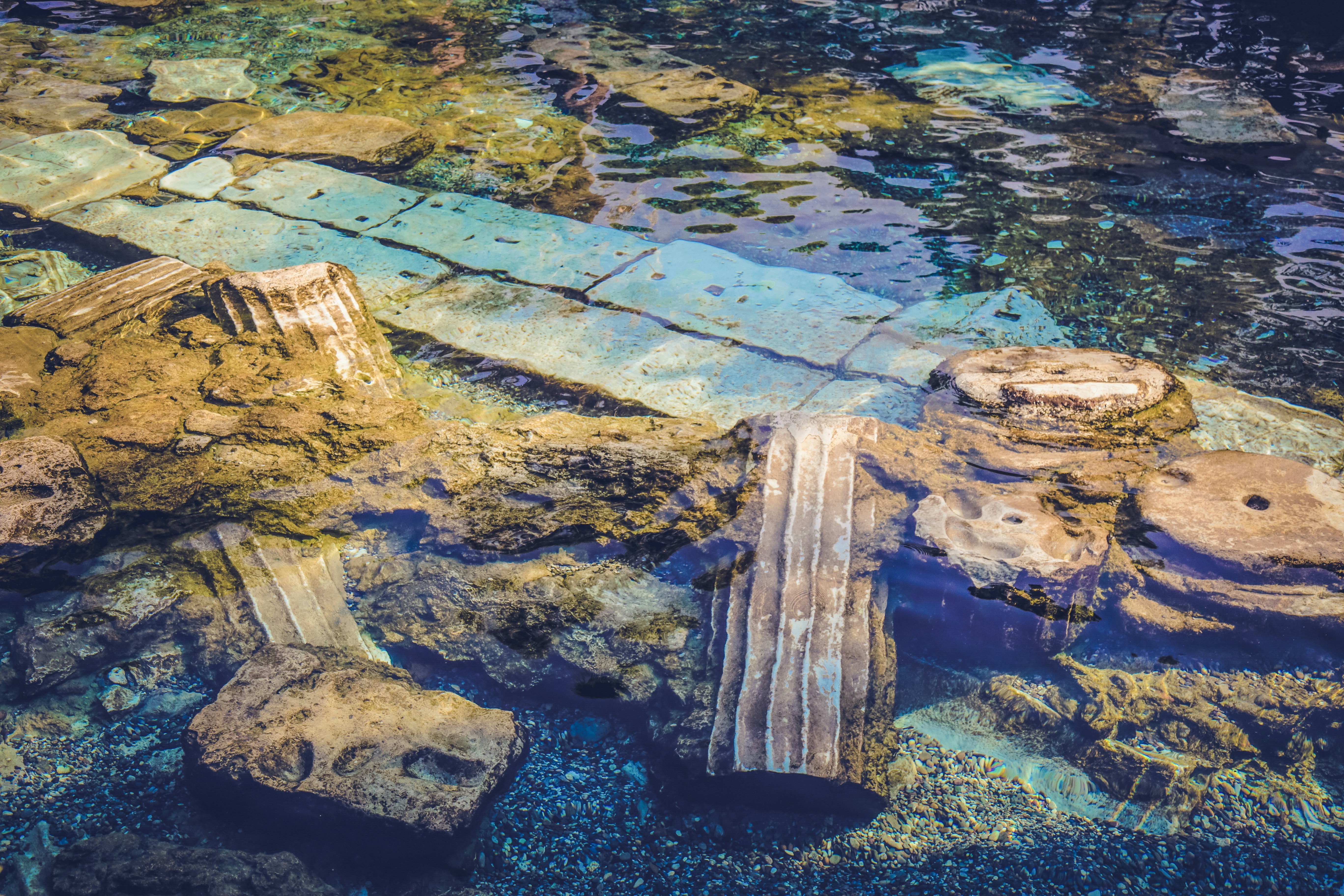 Cleared stone steps and columns submerged in vivid turquoise water, revealing an ancient, sunlit passage beneath a crystal pool. The image captures an underwater architectural detail with natural light playing on textures.