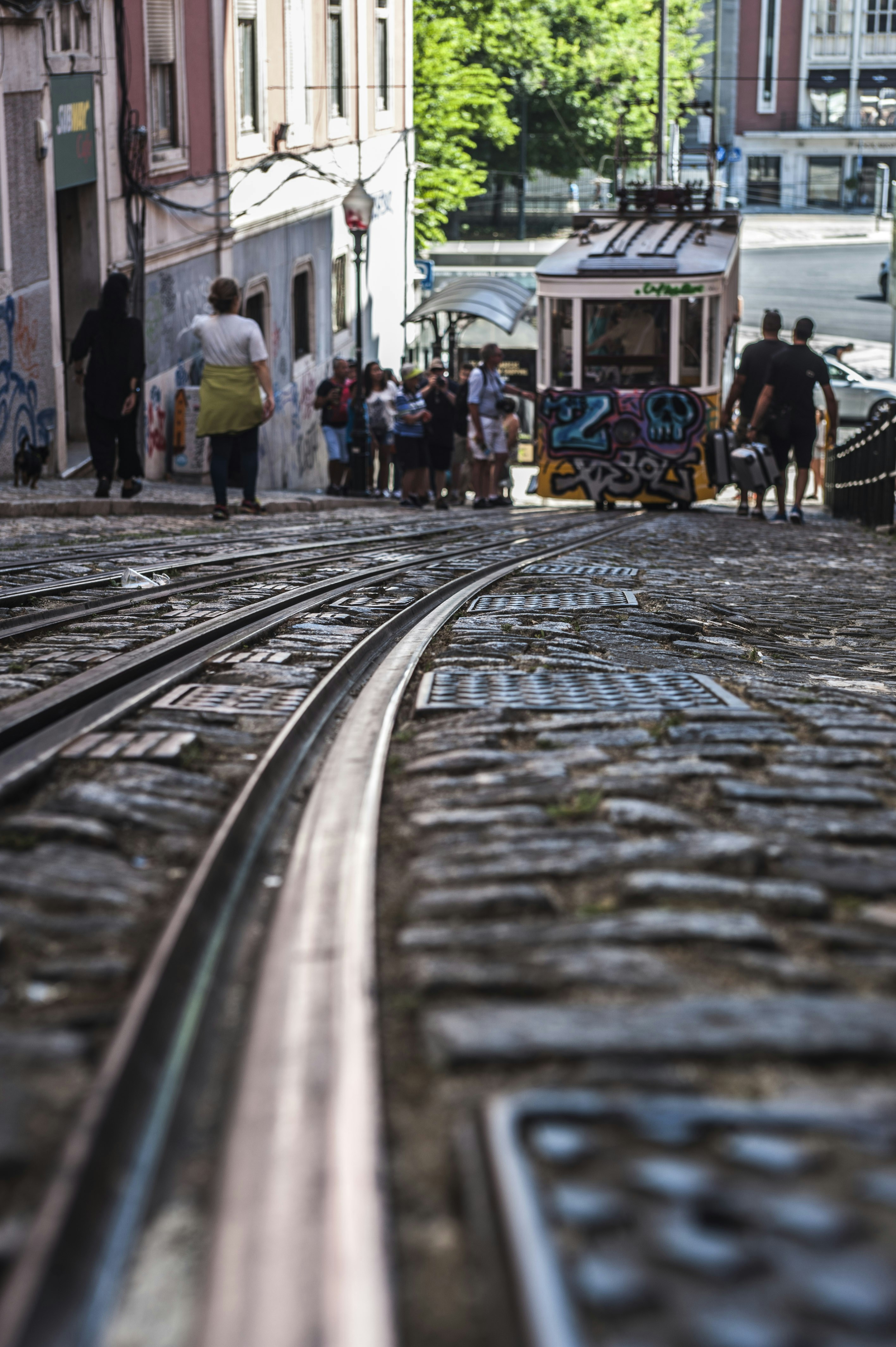 Cobblestone street with tram tracks curving toward a graffiti-covered tram stop; pedestrians gather near colorful walls and a small urban scene; a city tram sits in the background.