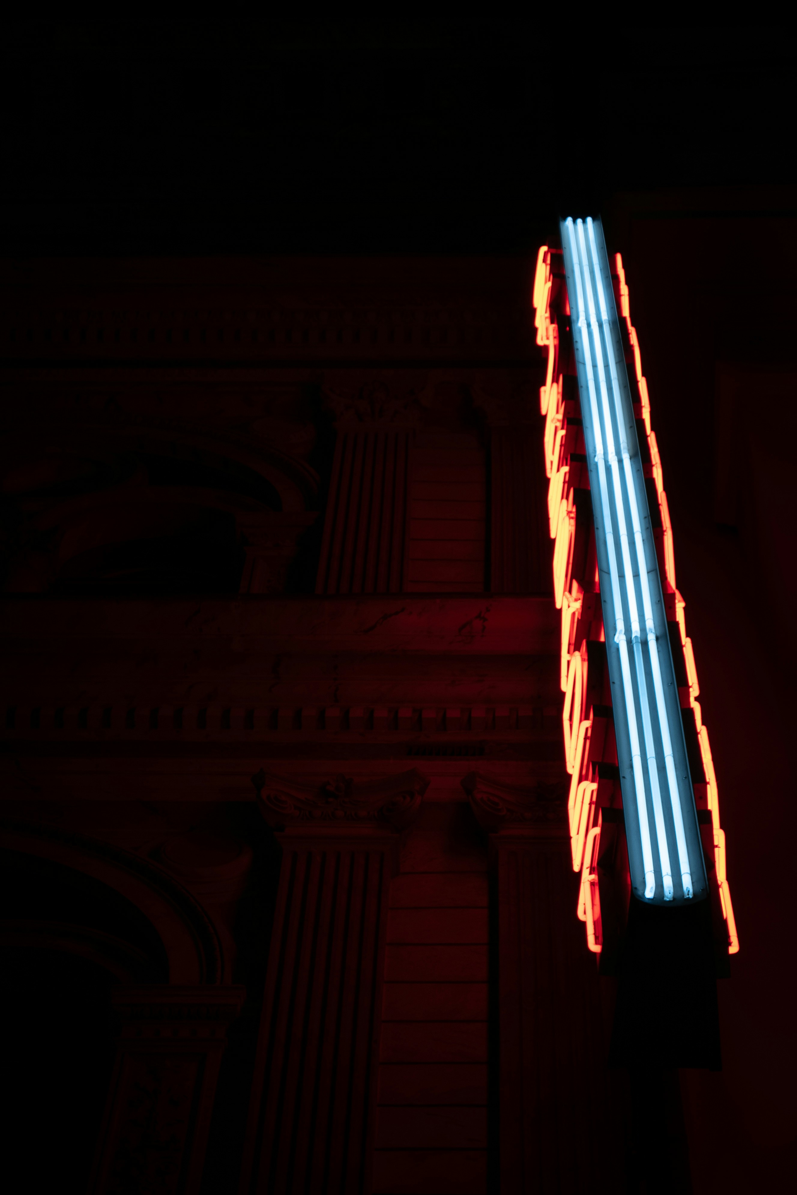 Vertical neon sign with blue tubes outlined in red against a dark historic building façade.