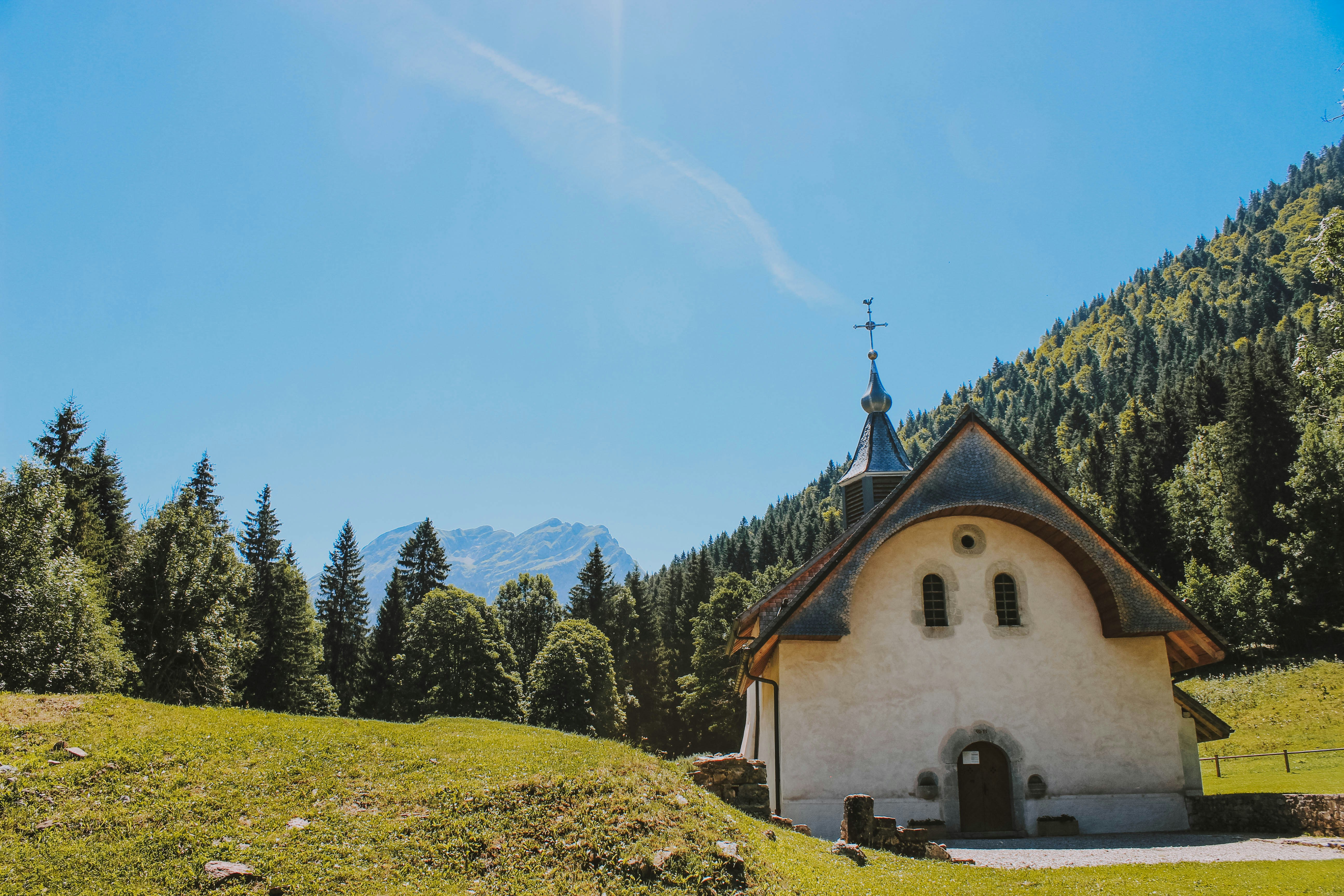 Charming chapel nestled in a lush green valley, framed by majestic mountains under a clear blue sky.