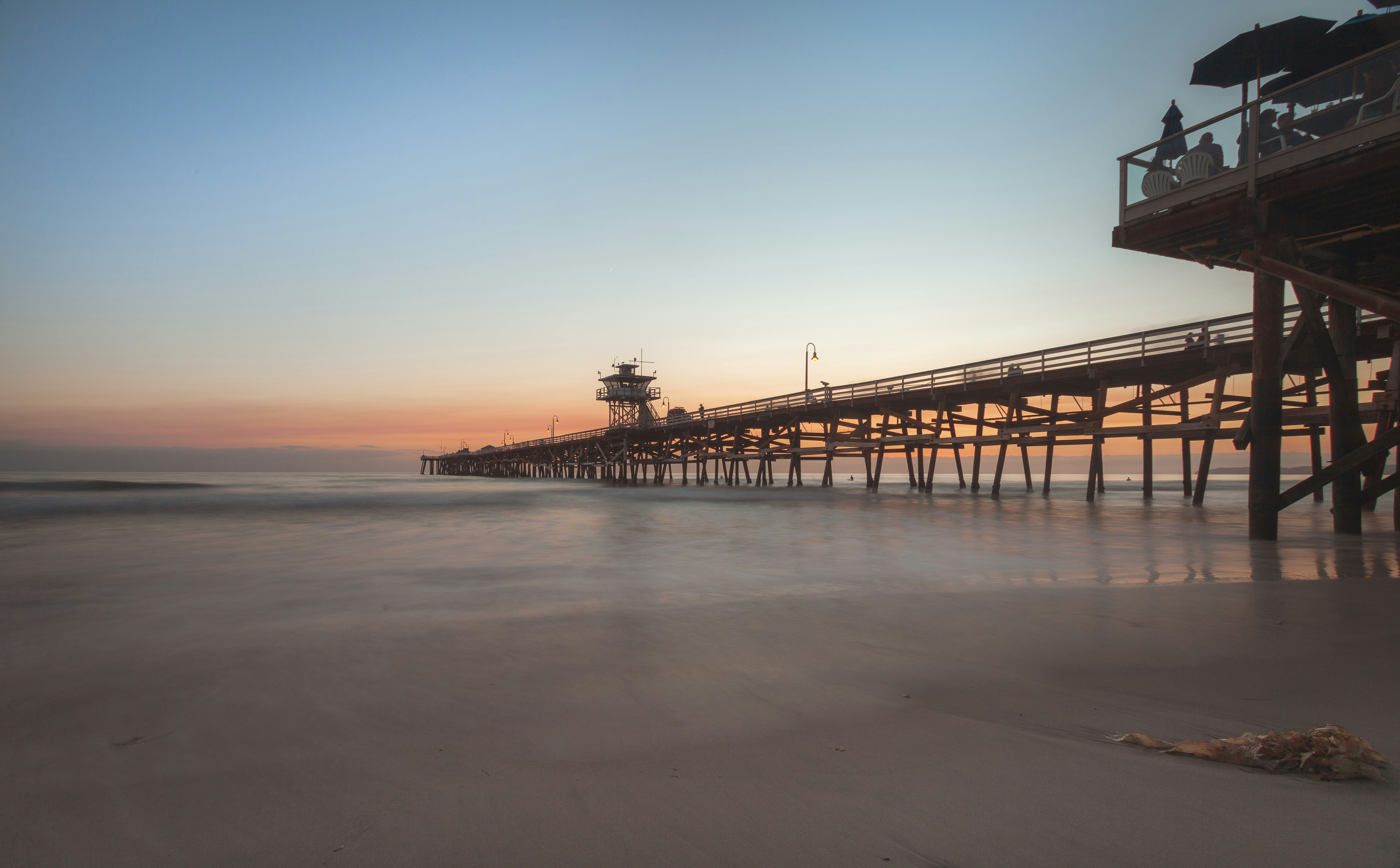 Long wooden pier stretching into calm ocean waters at sunset with soft pastel skies.