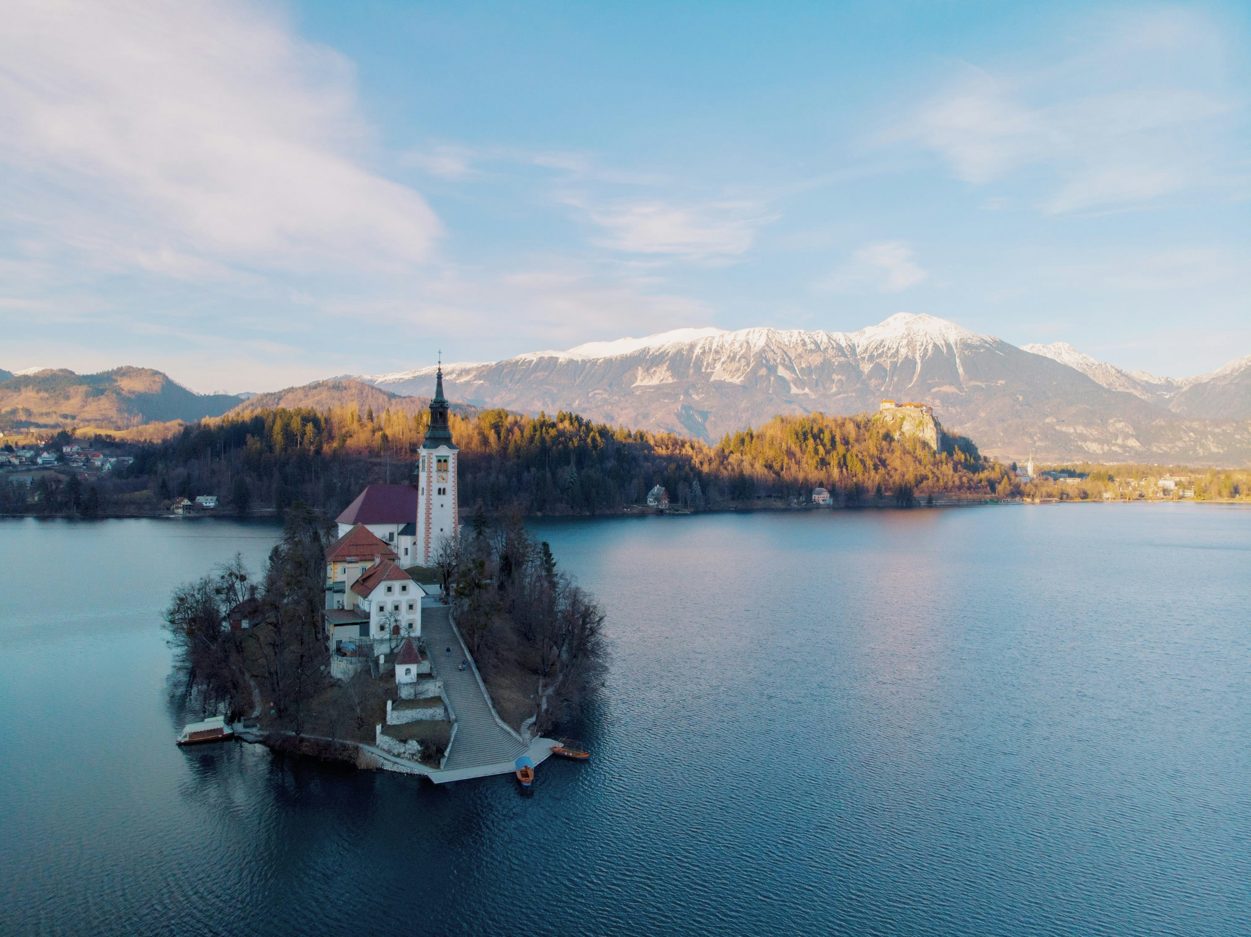 Aerial view of Bled Island with a church surrounded by a calm lake and snow-capped mountains under a clear blue sky.