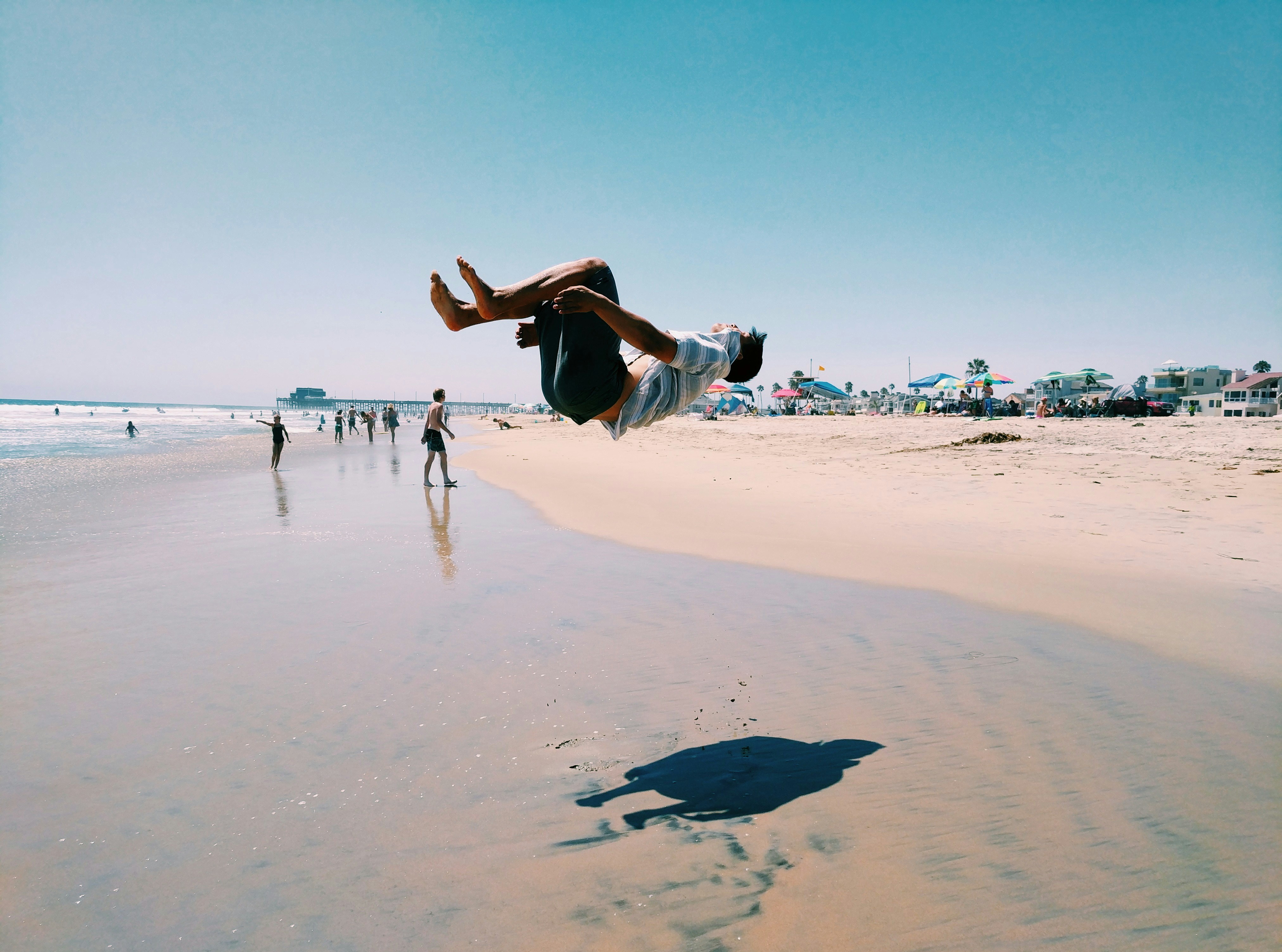 A person performing a backflip above the water's edge, with beachgoers and colorful umbrellas in the background.
