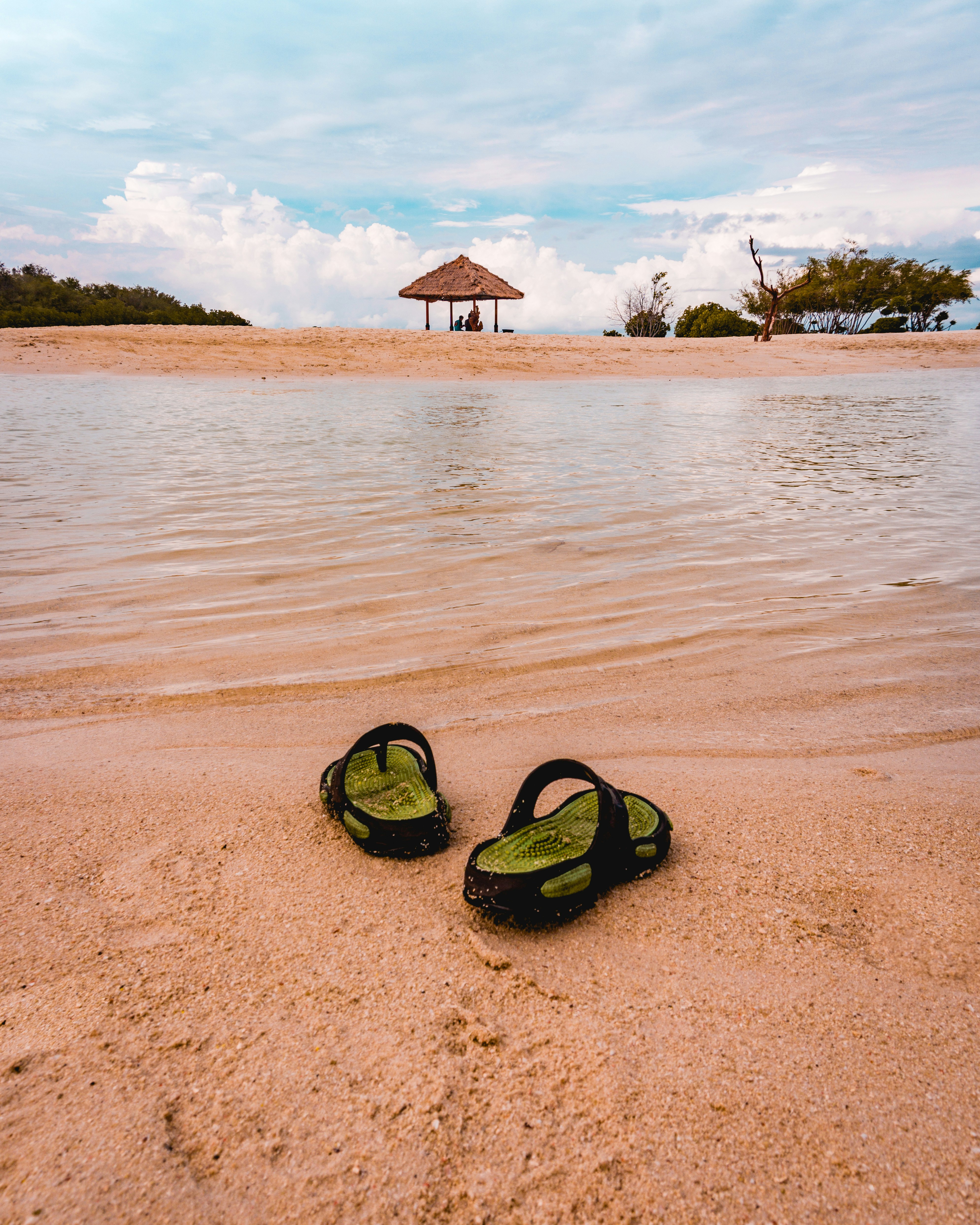 Sand beach with a pair of green flip-flops in the foreground and a thatched hut near the waterline under a soft sky; a calm, tropical idle scene.