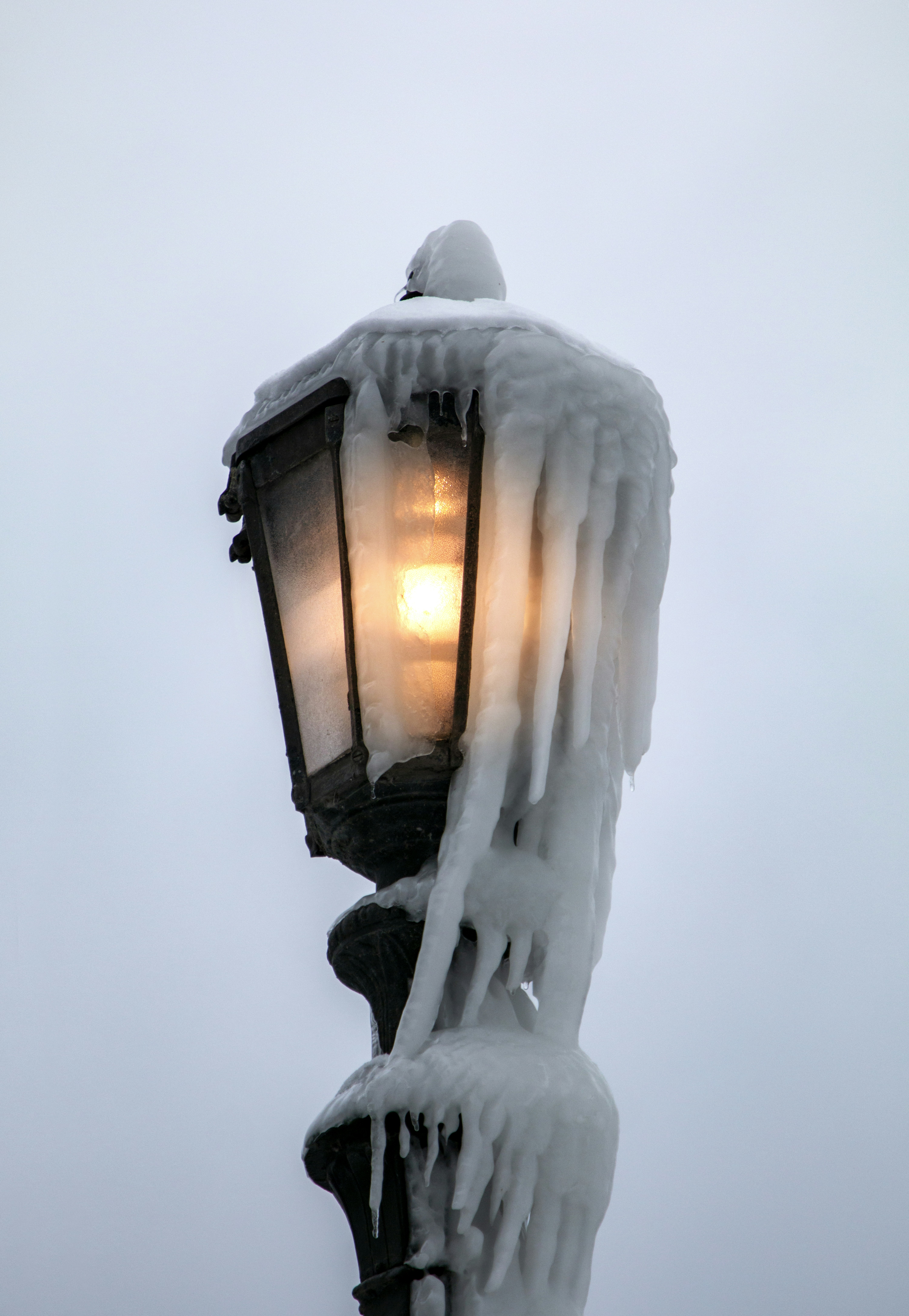 Icicles cling to an ornate street lamp, illuminating the wintry scene with a warm glow against a gray sky.