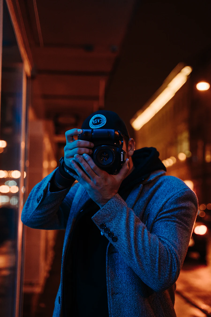 Person in a blue coat and cap holding a Canon camera up to their face on a warmly lit city street at night. Blurred lights and reflections create a moody urban background.