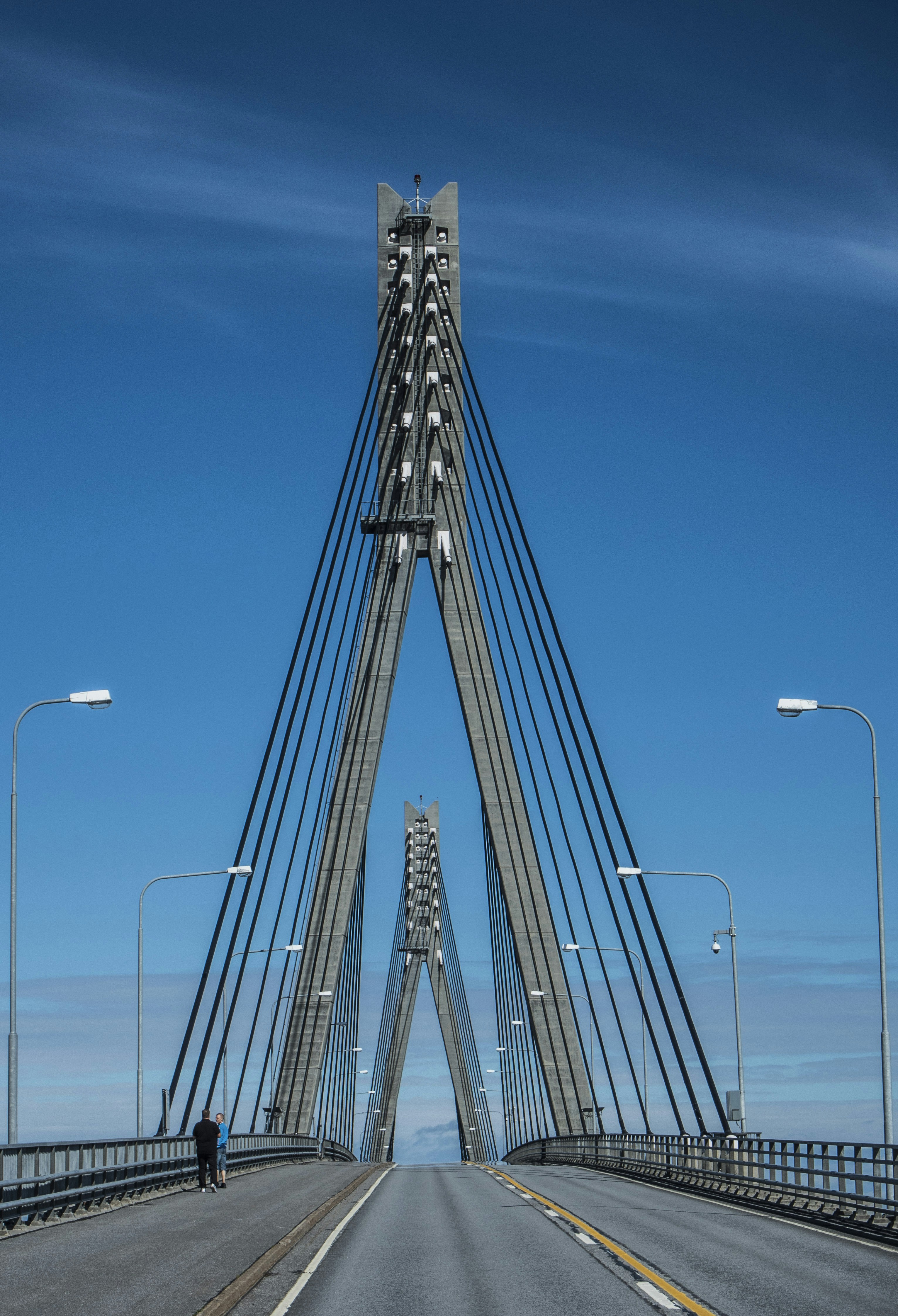Photograph of a cable-stayed bridge with a central tower and converging cables against a vivid blue sky; a solitary figure walks the deck.