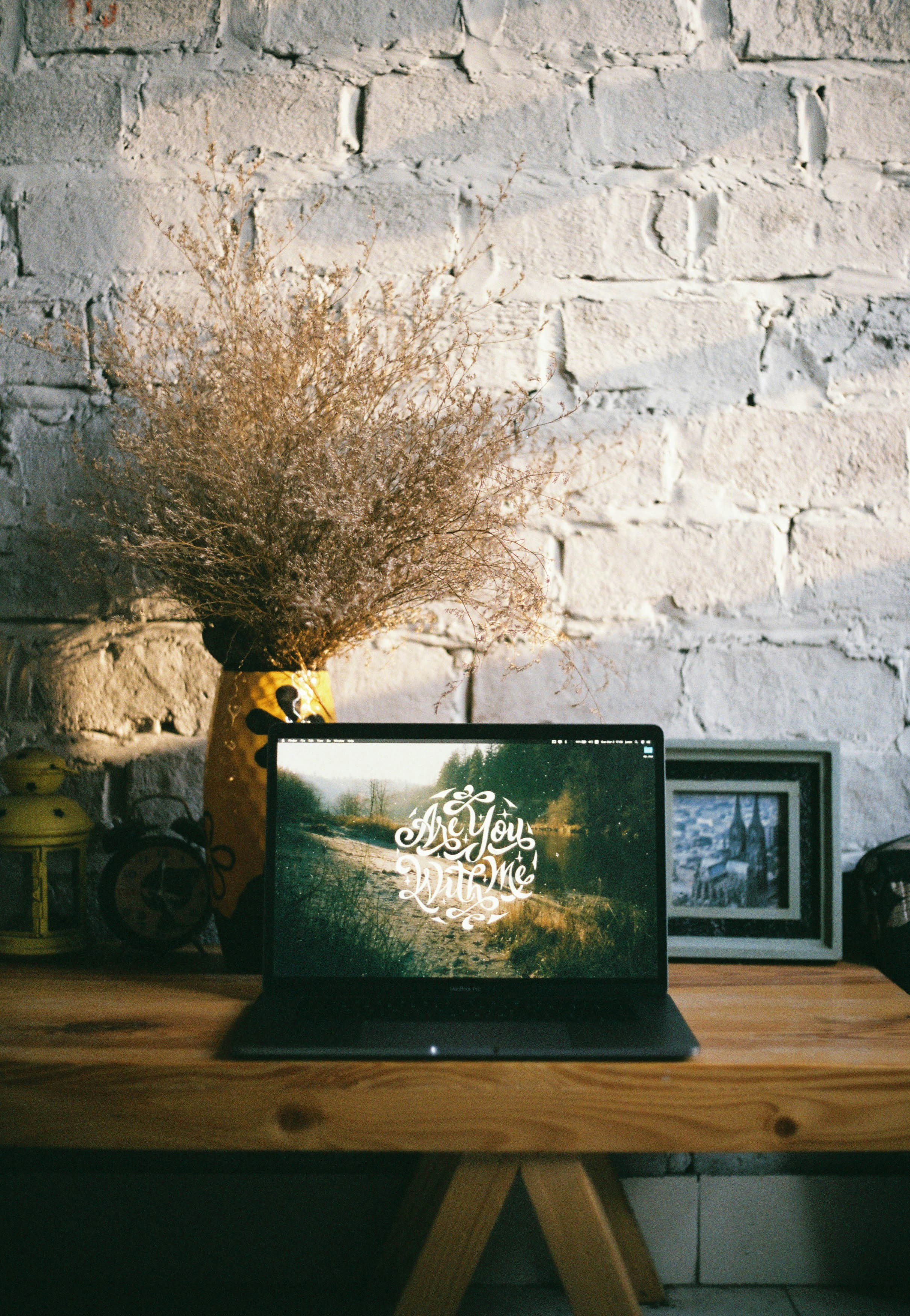 A laptop displaying a scenic landscape sits on a wooden table, complemented by a vase of dried flowers against a textured white brick wall.