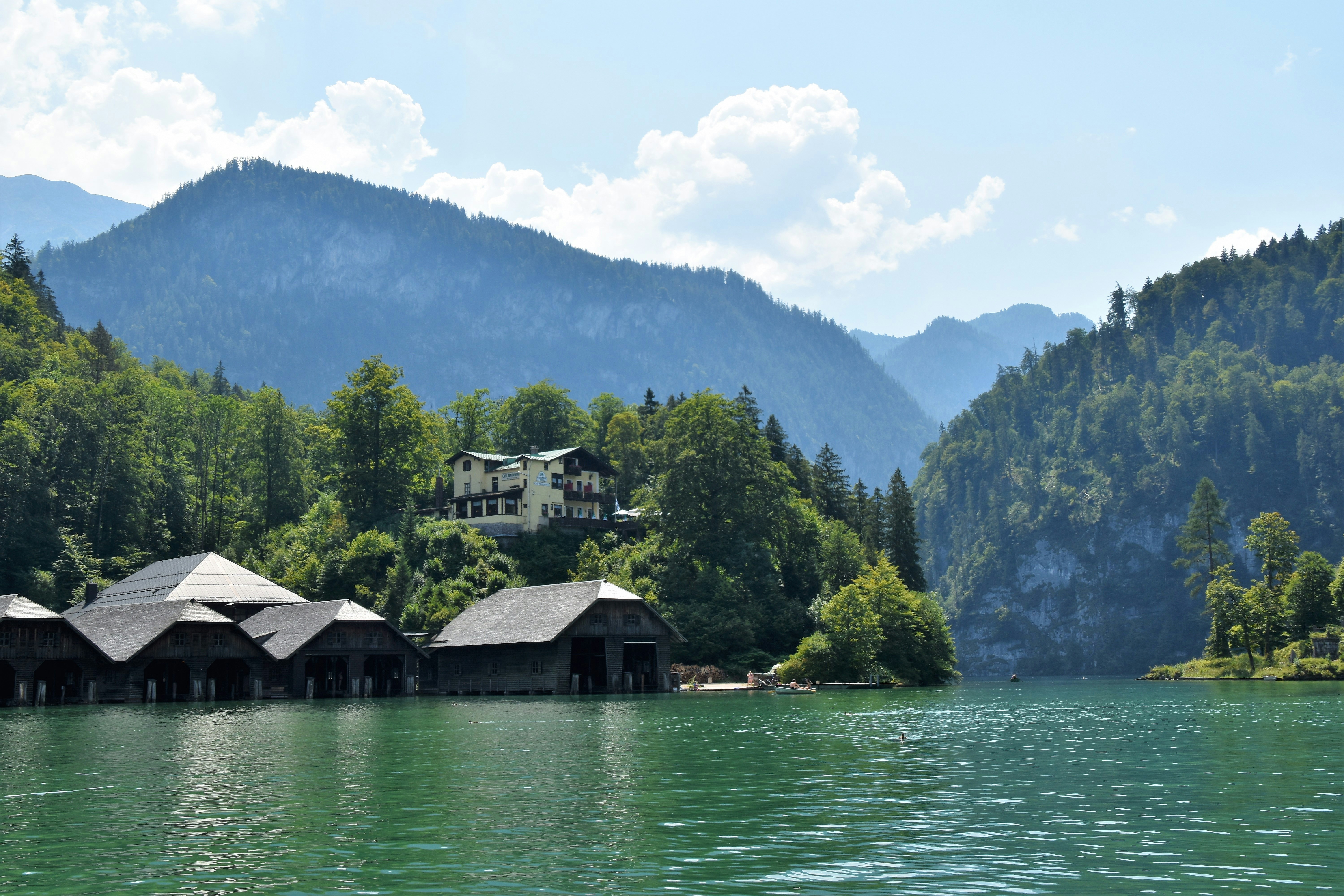 Lakeside boathouses reflect in calm waters, framed by lush greenery and towering mountains under a vibrant sky.