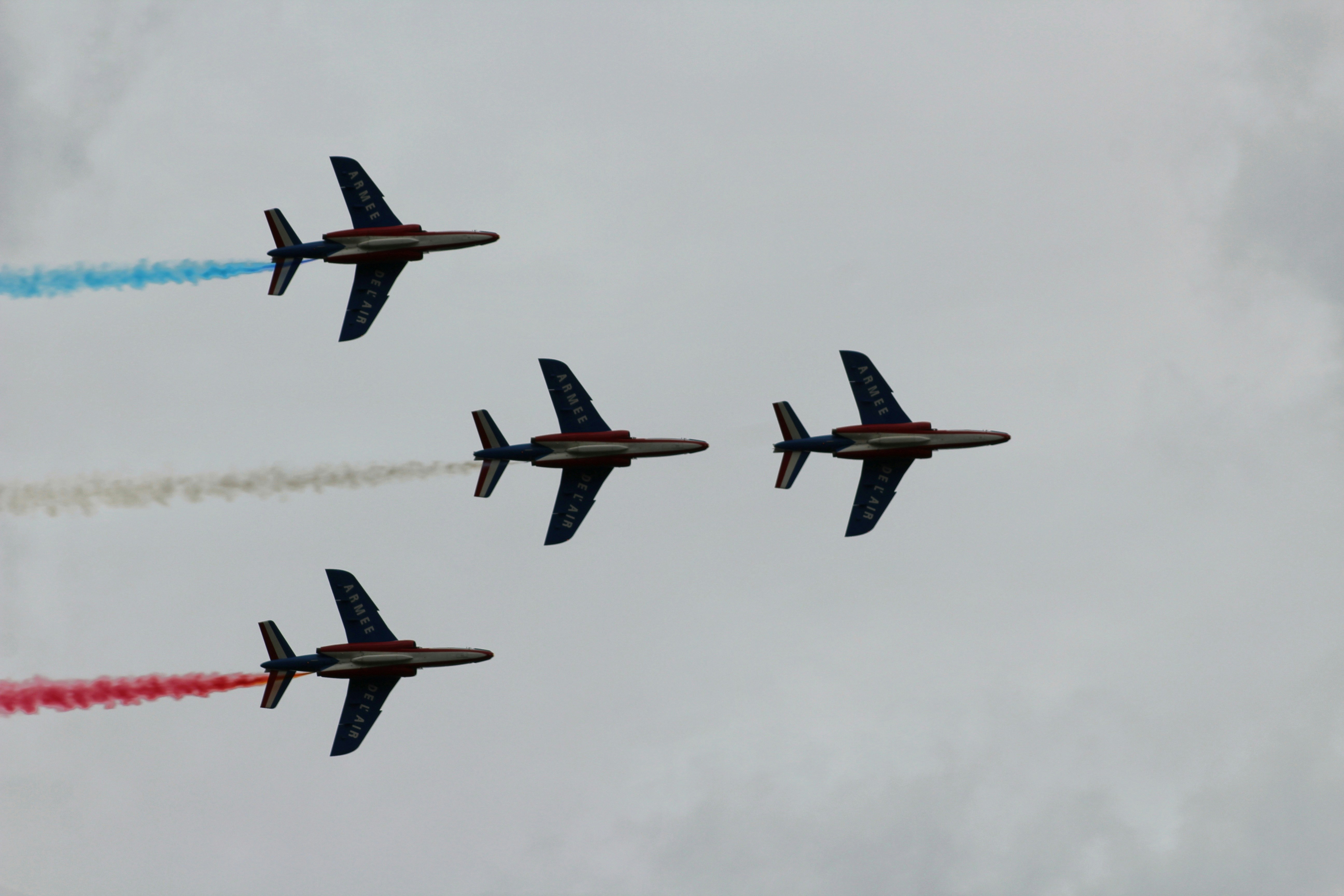 Four fighter jets performing a synchronized maneuver, trailing colored smoke in red, white, and blue against a cloudy sky.