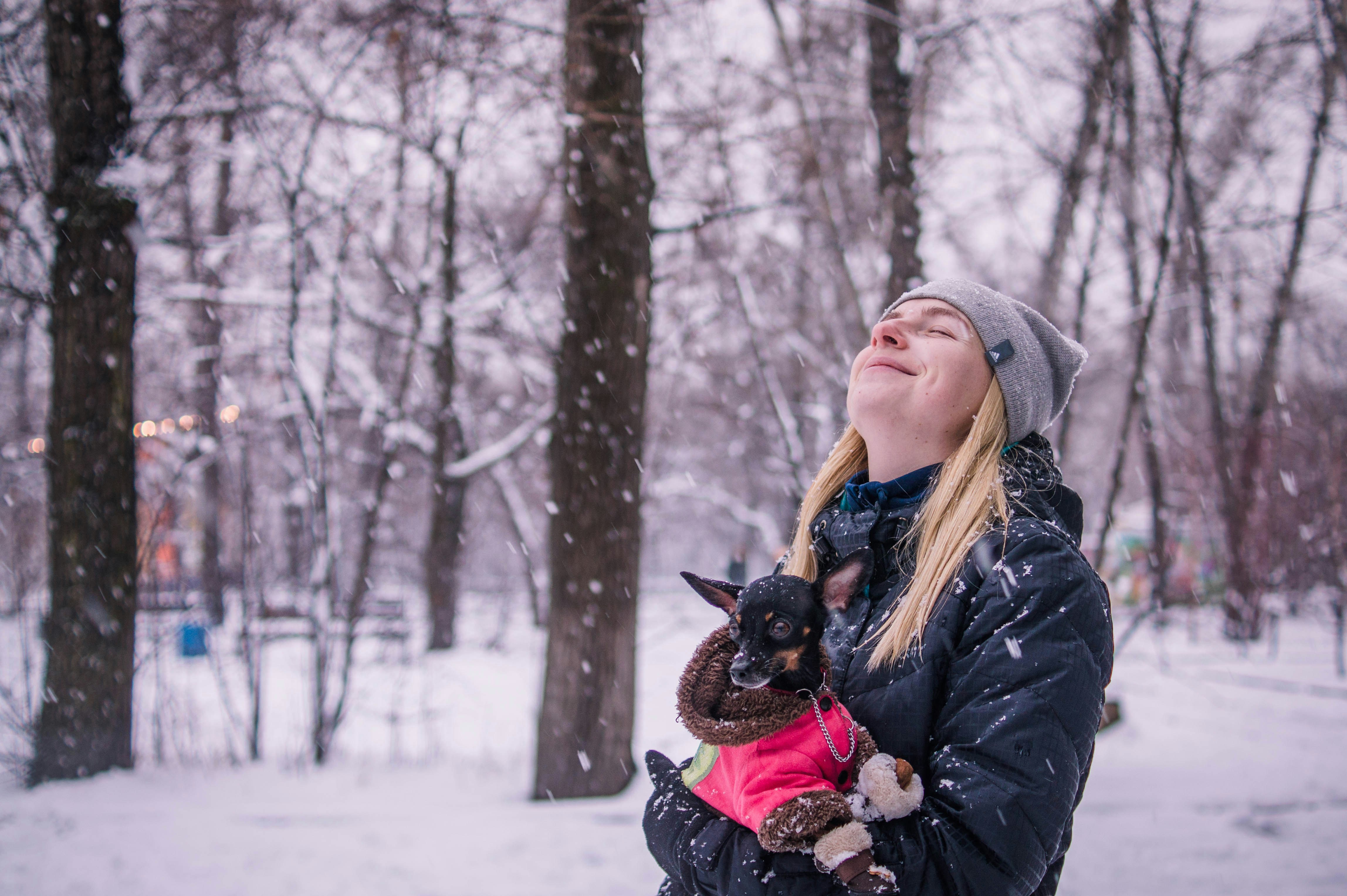 Woman holding a small dog in a pink coat, standing amidst snow-covered trees, gazing upwards with a serene expression.