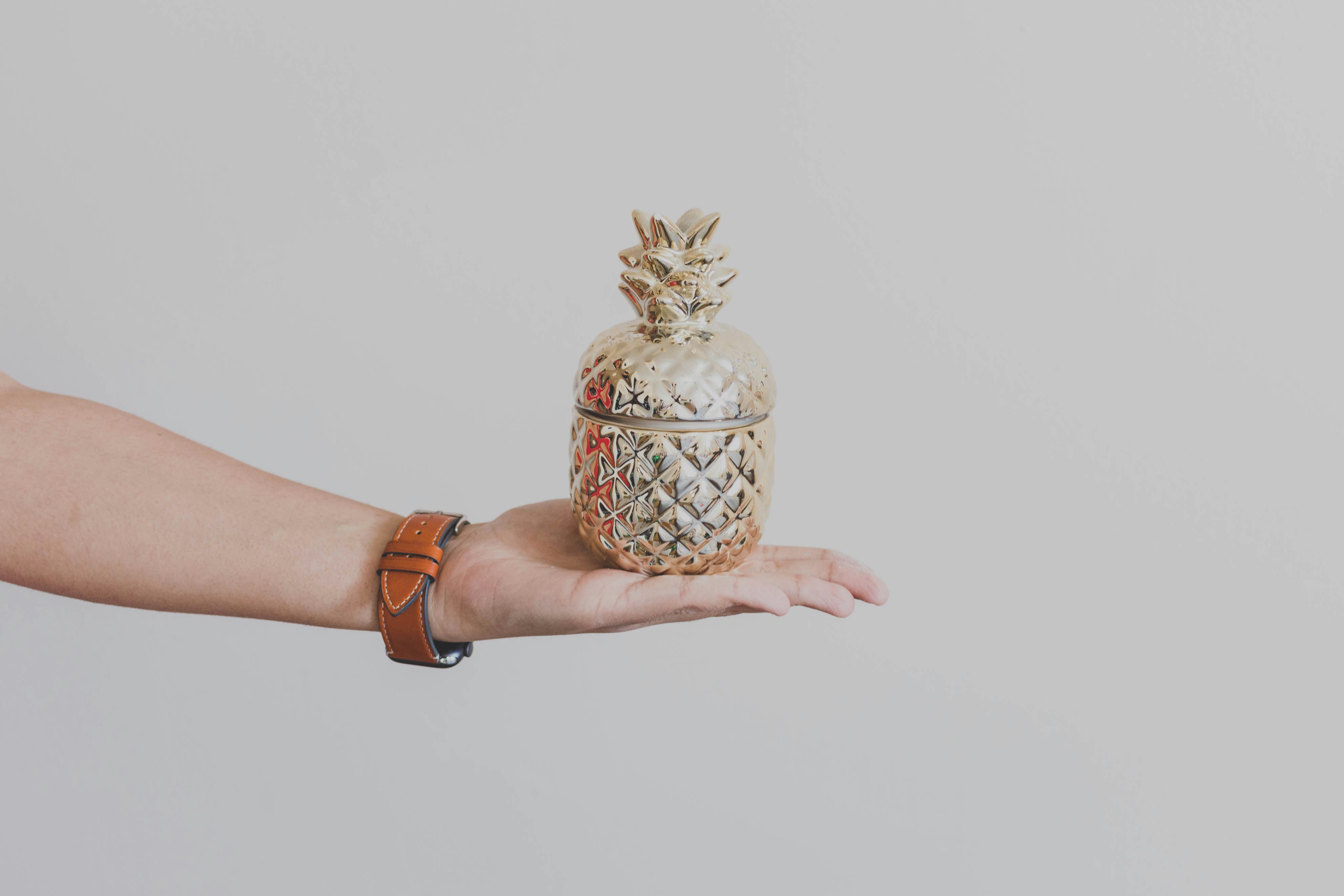 Hand holding a decorative golden pineapple container against a neutral background.