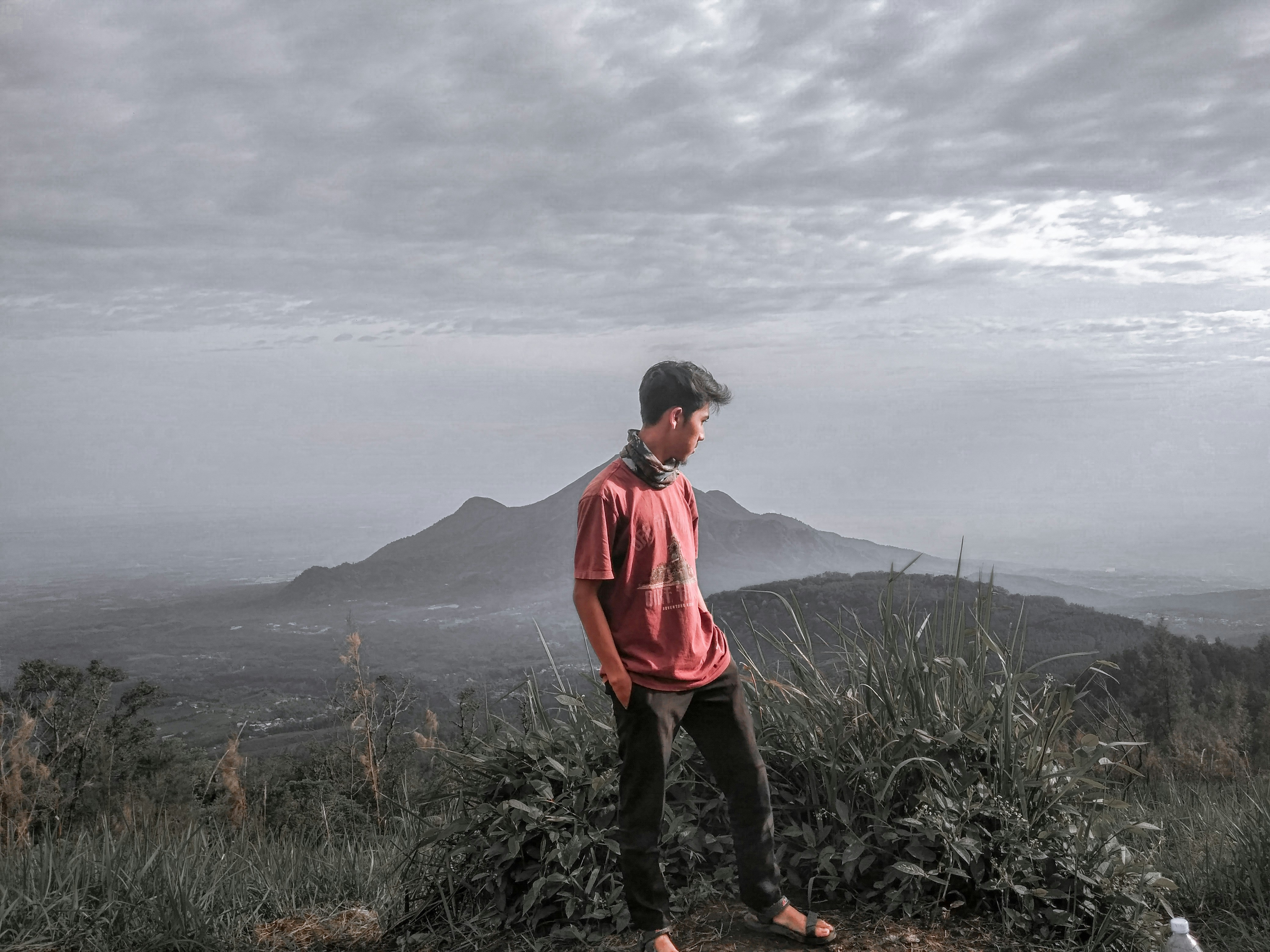 Young man standing on a hillside, gazing at distant mountains under a cloudy sky. The scene captures a serene connection with nature.