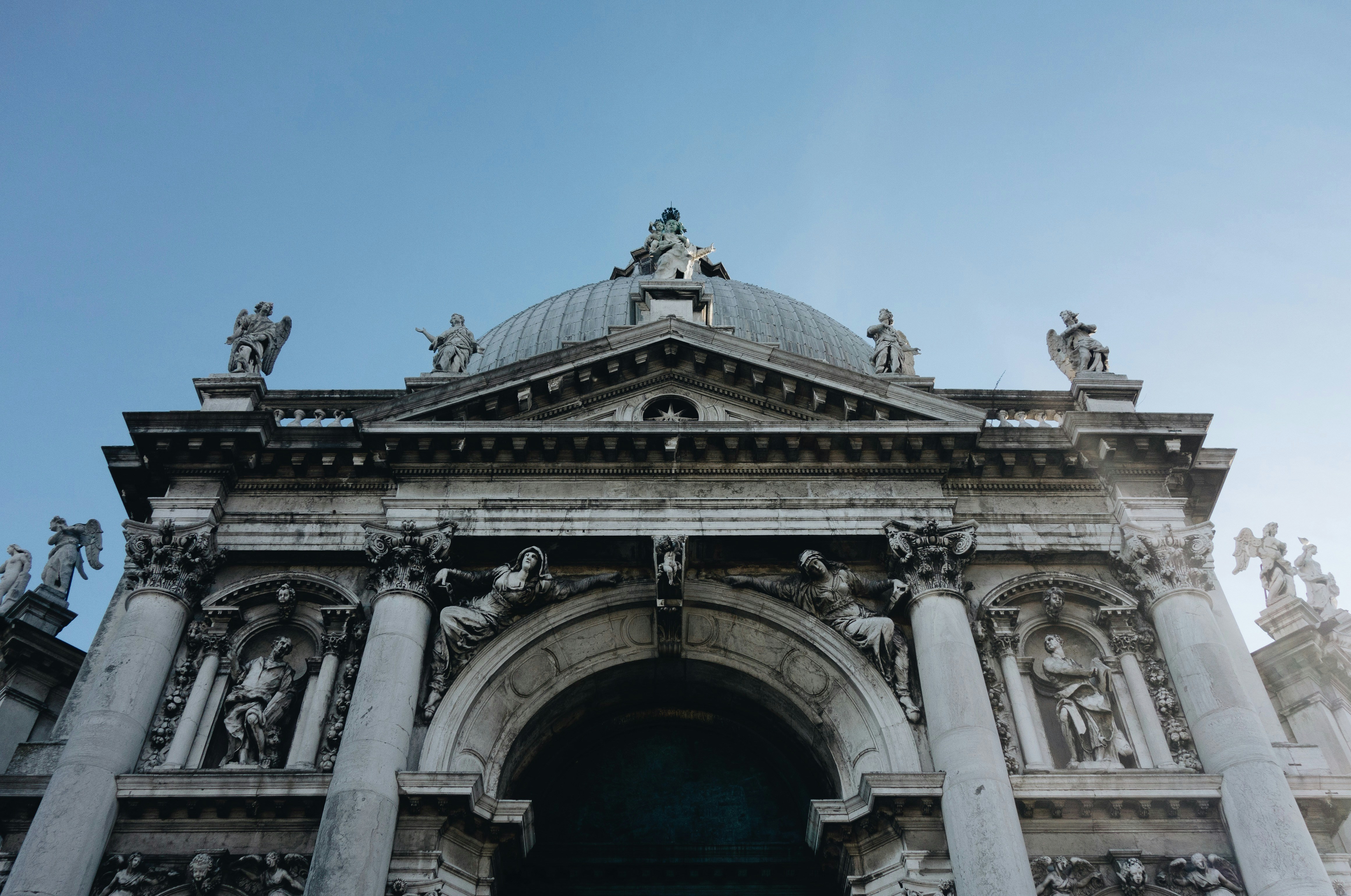The image captures the grand facade of a historic building adorned with intricate sculptures and towering columns, set against a clear blue sky. The soft lighting highlights the detailed stonework and creates a serene atmosphere, emphasizing the architectural elegance. This striking composition draws attention to the symmetrical beauty and the imposing presence of the structure.