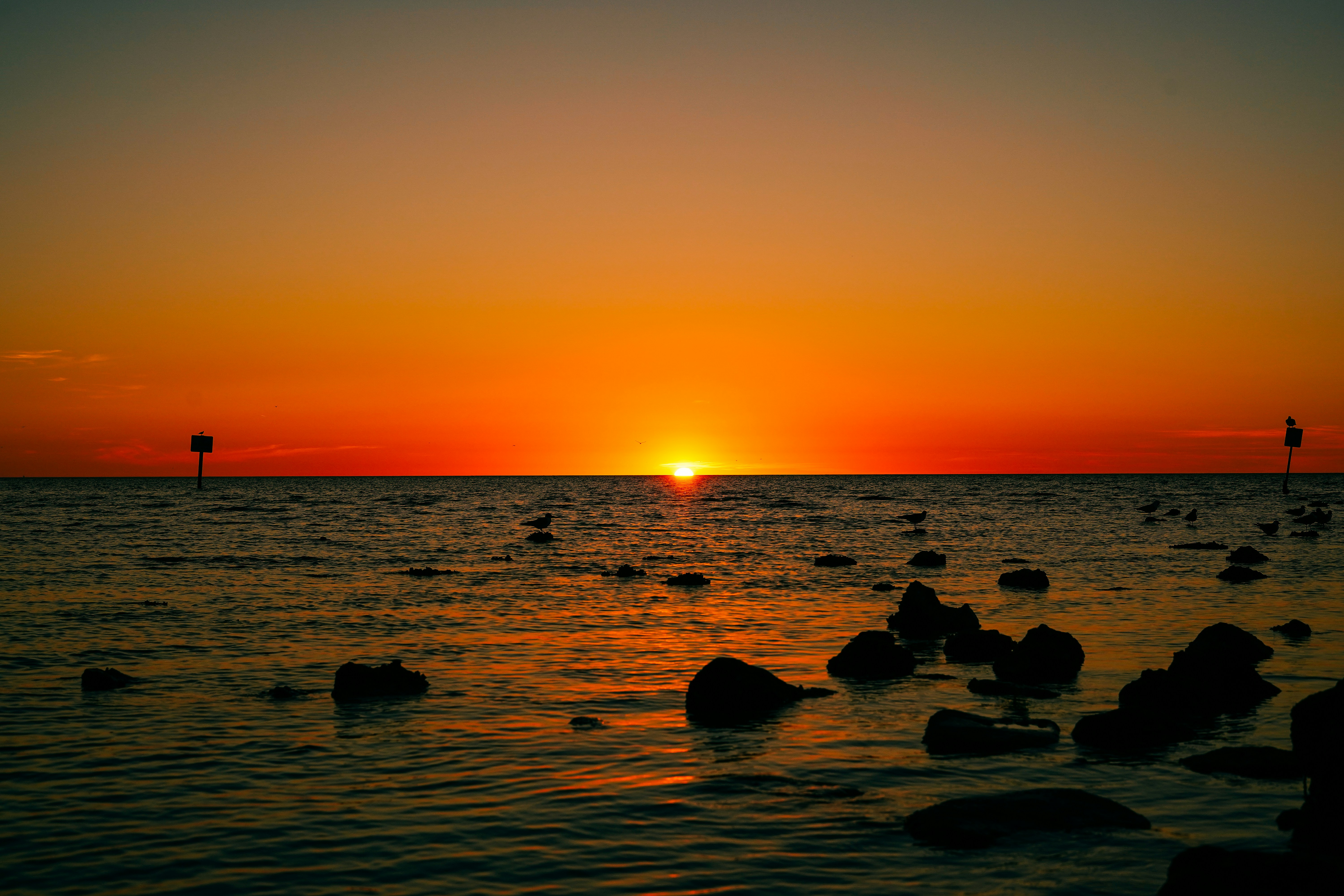 Golden sun setting over a calm ocean with scattered rocks and vibrant sky hues.