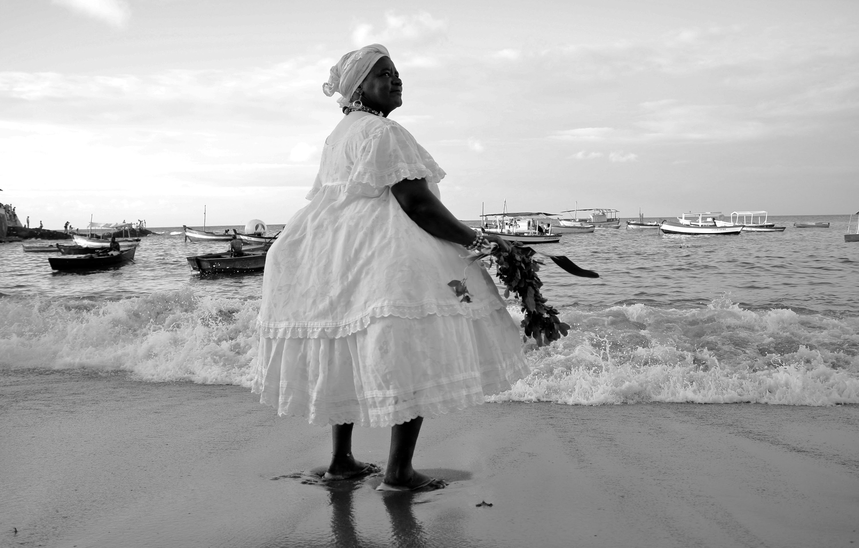 Woman in traditional dress stands at the water's edge holding flowers, with boats in the background under a cloudy sky.