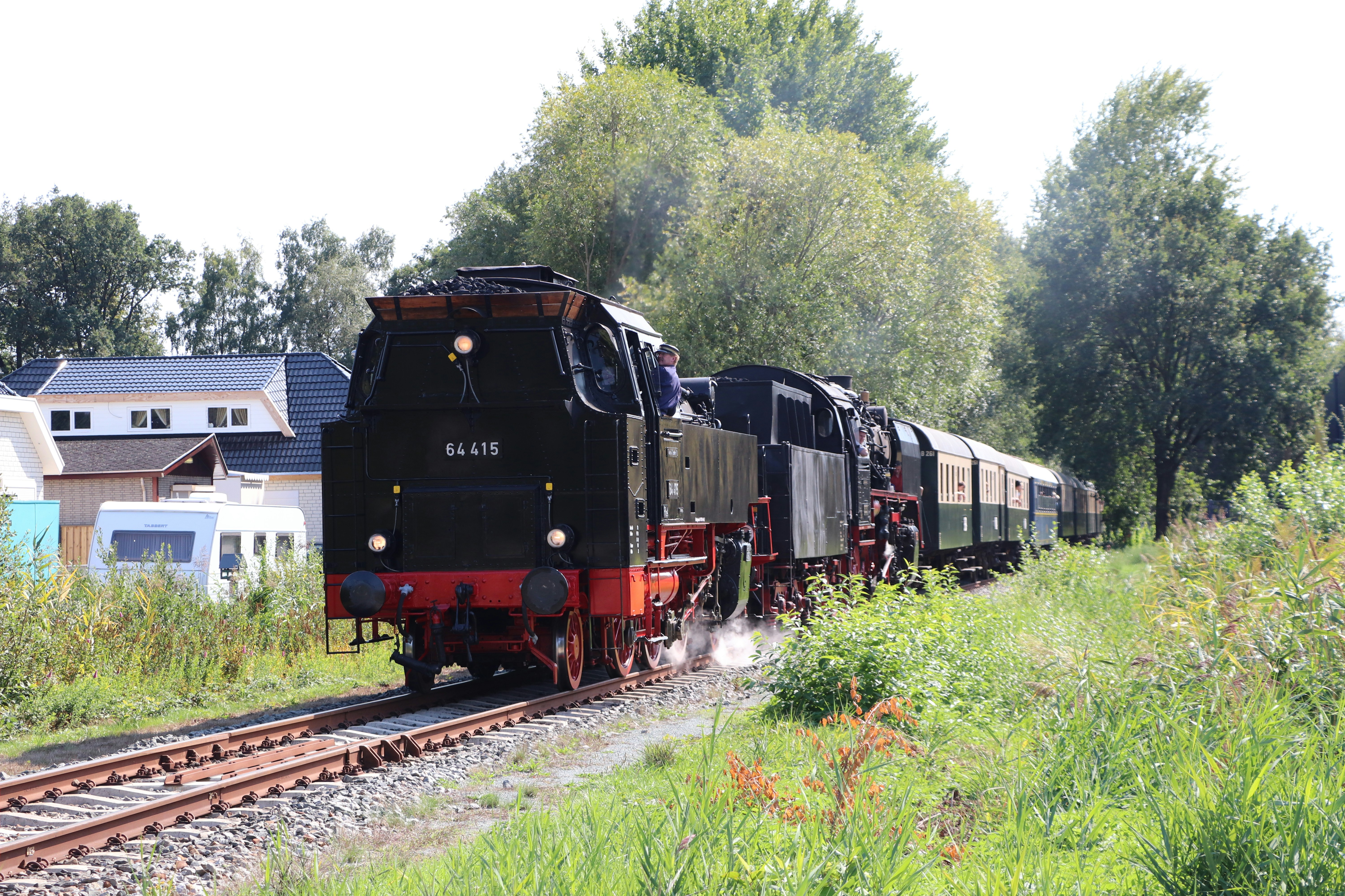 Steam locomotive pulling vintage carriages along a rural track, surrounded by lush greenery and distant houses under a clear sky.
