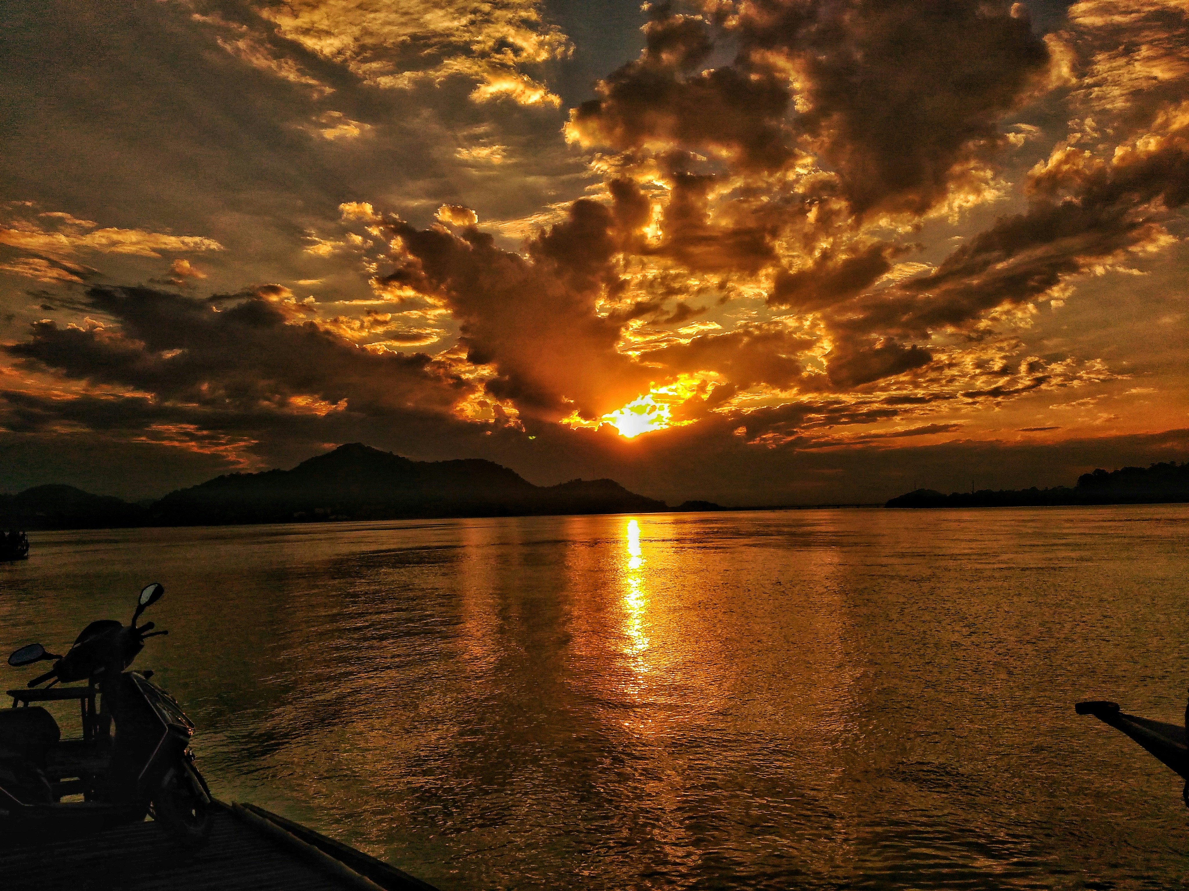 Dramatic sunset over a calm lake with silhouetted mountains and a motorbike on the shore.