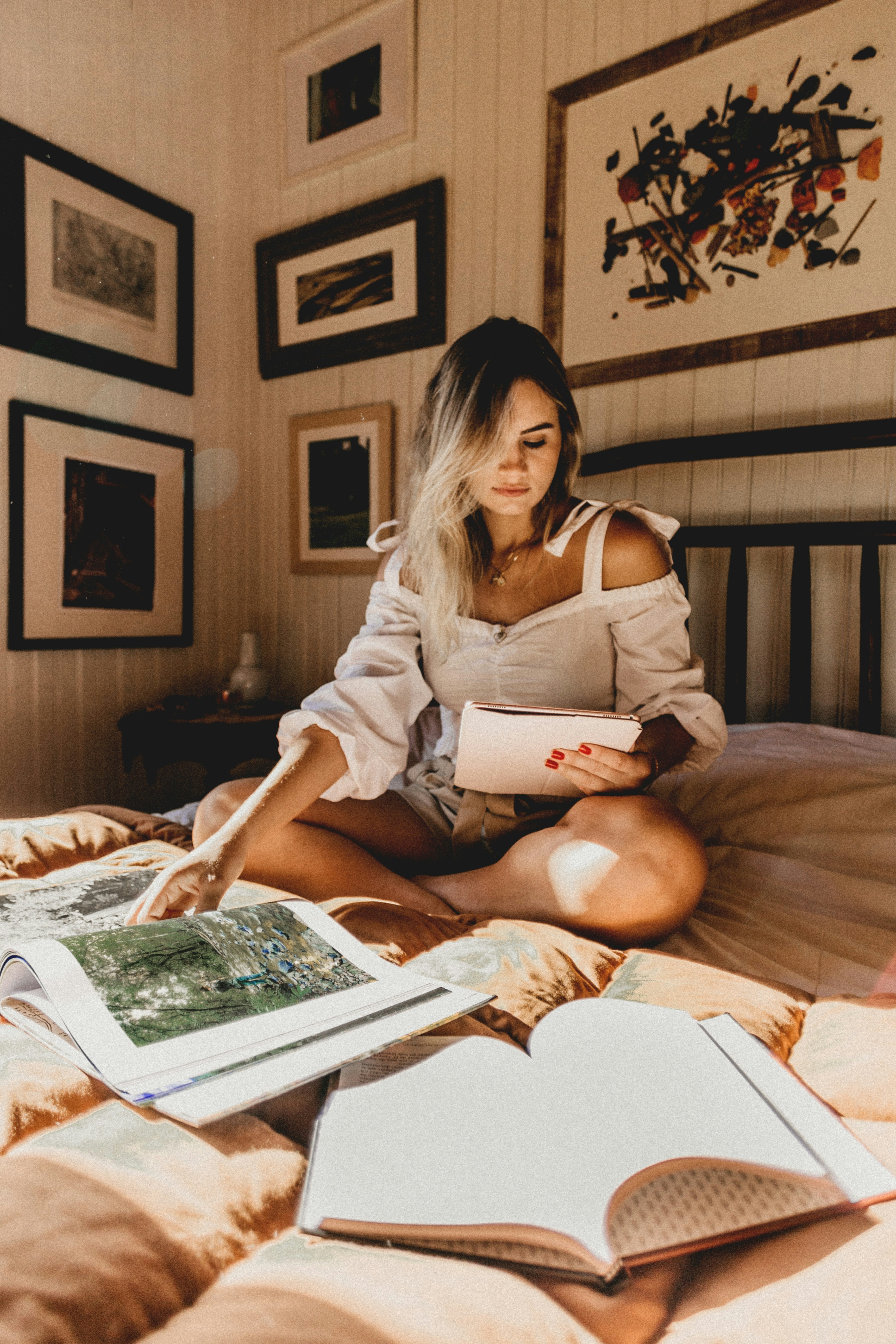 Young woman seated on a bed, examining an open book while surrounded by framed art in a sunlit room.