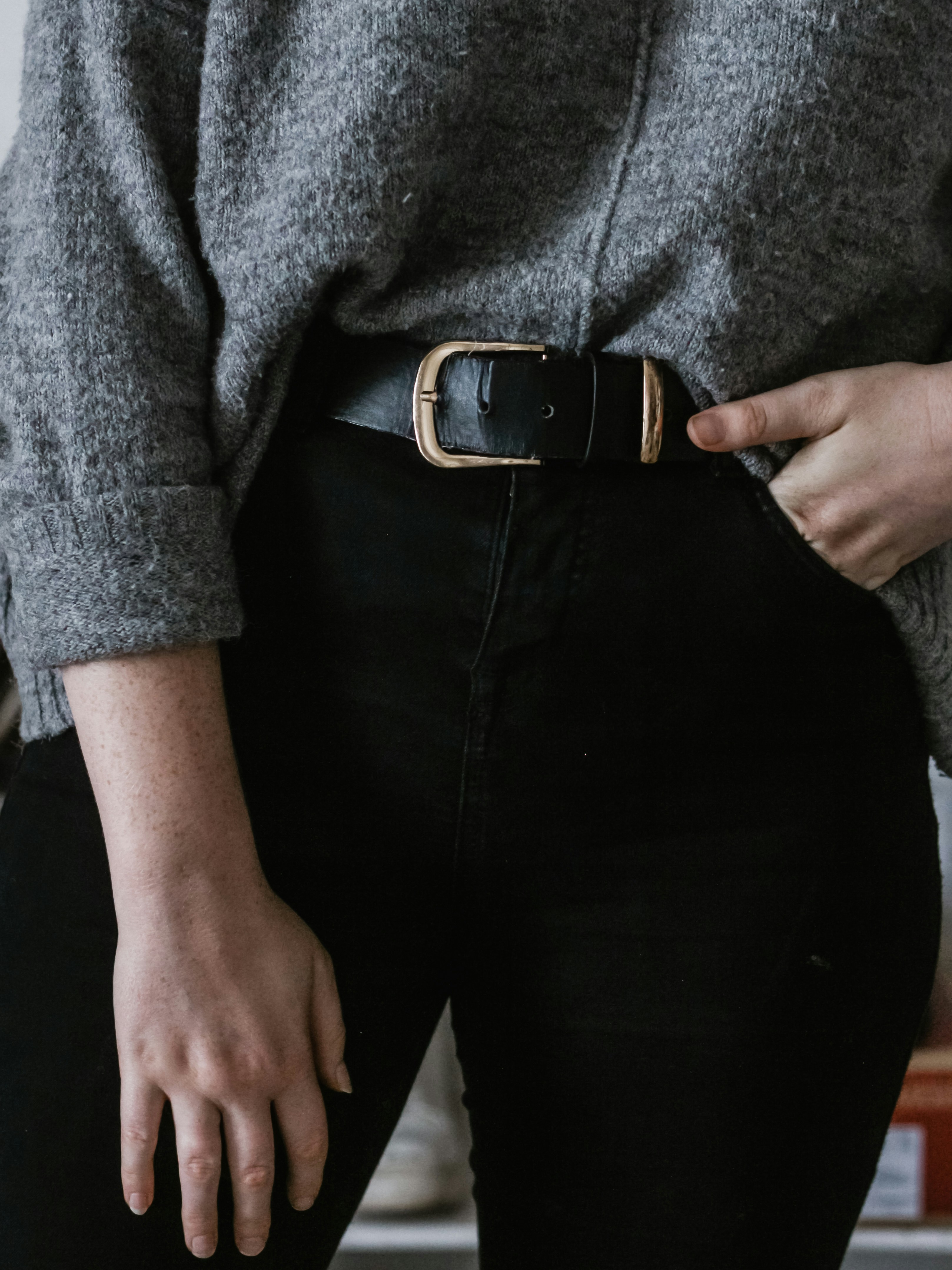 Close-up of a person wearing a cozy gray sweater and black jeans, with a focus on their stylish belt. The outfit combines comfort and fashion seamlessly.