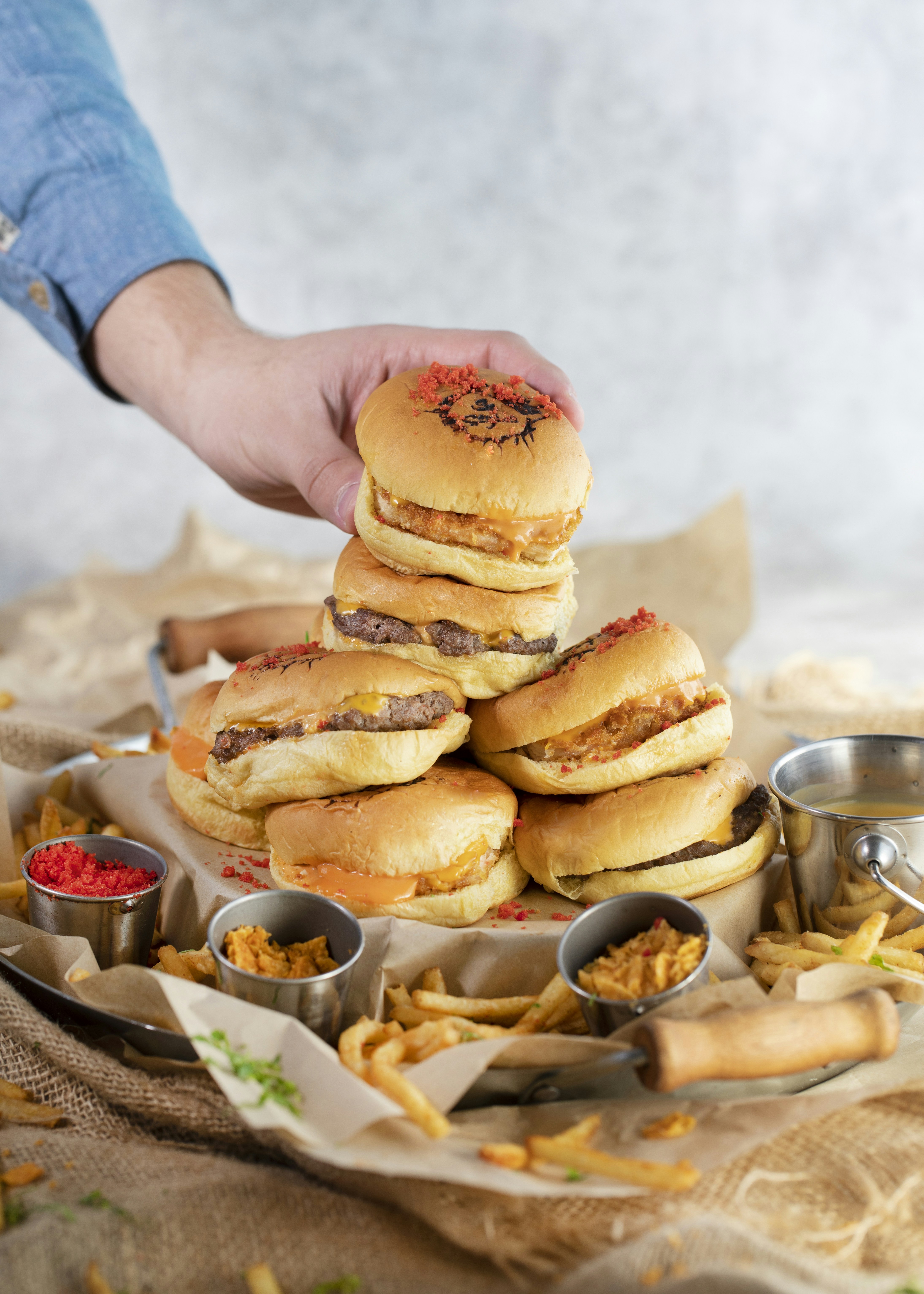 A hand reaches for a towering stack of gourmet burgers, surrounded by crispy fries and colorful toppings, showcasing a delightful culinary arrangement.