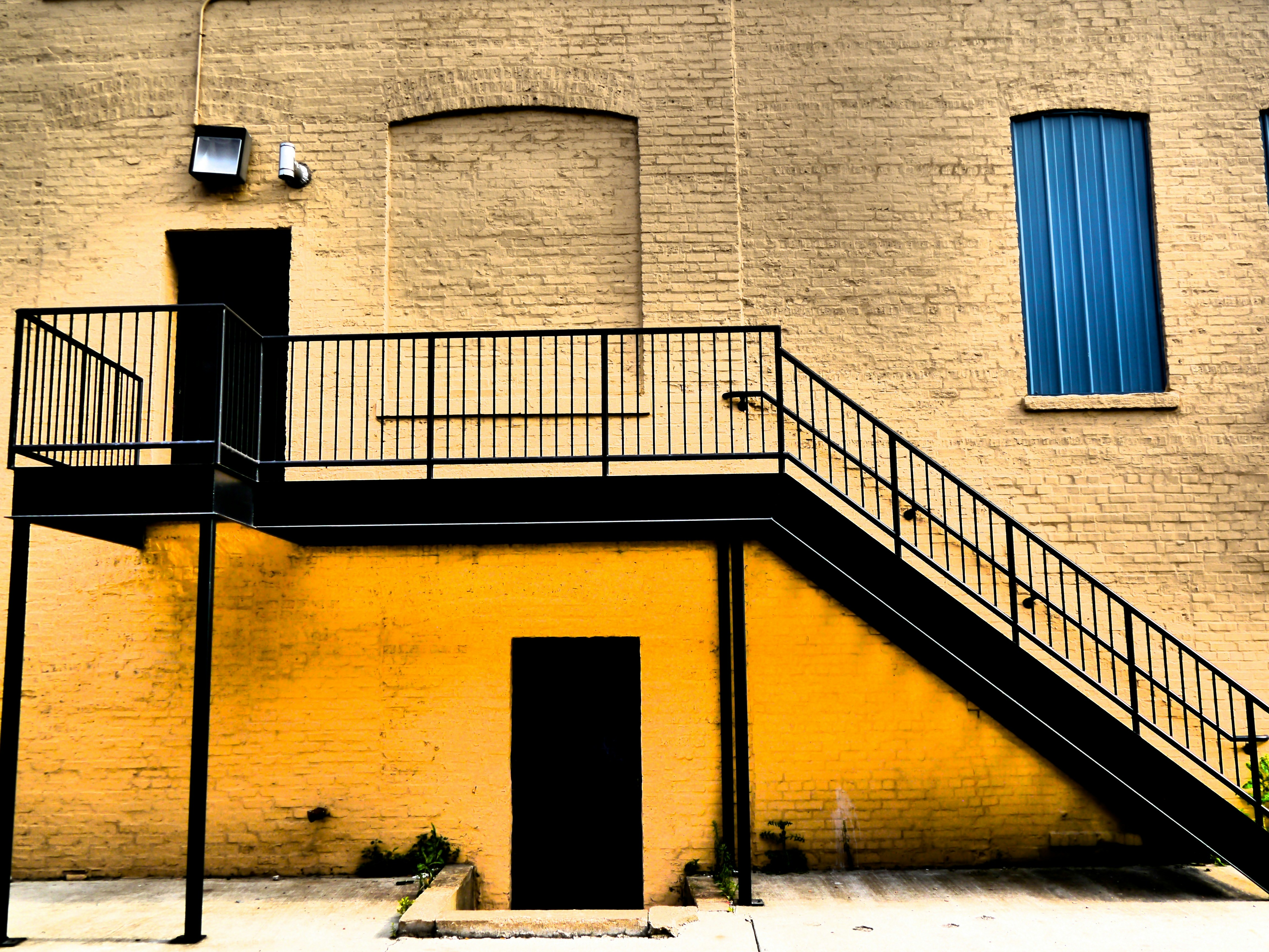 Industrial staircase leading up to a door against a textured yellow wall, with contrasting blue window above. 