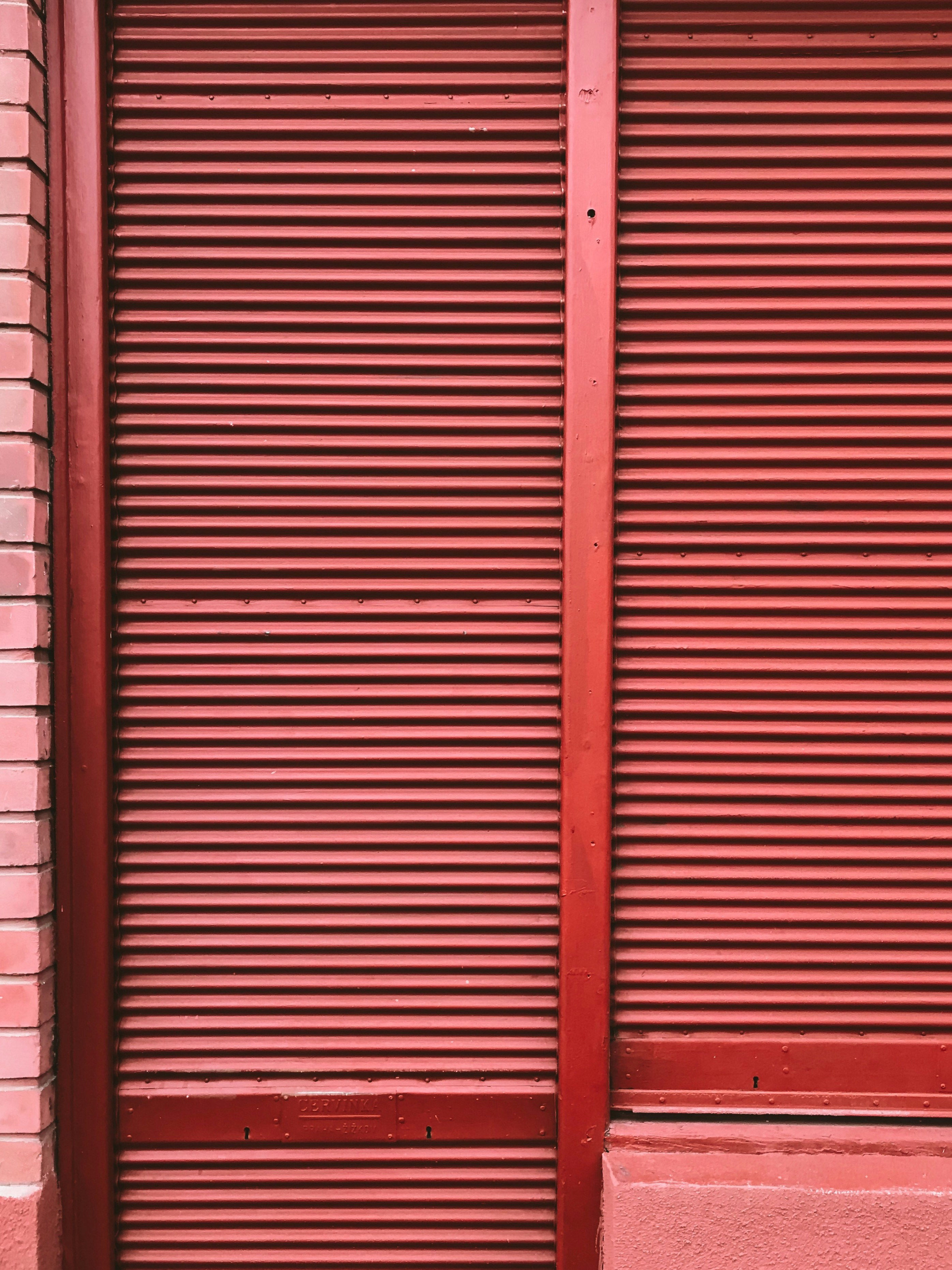 Textured red metal shutter with horizontal lines, complemented by a brick wall on the left side. The image highlights the interplay of color and pattern.