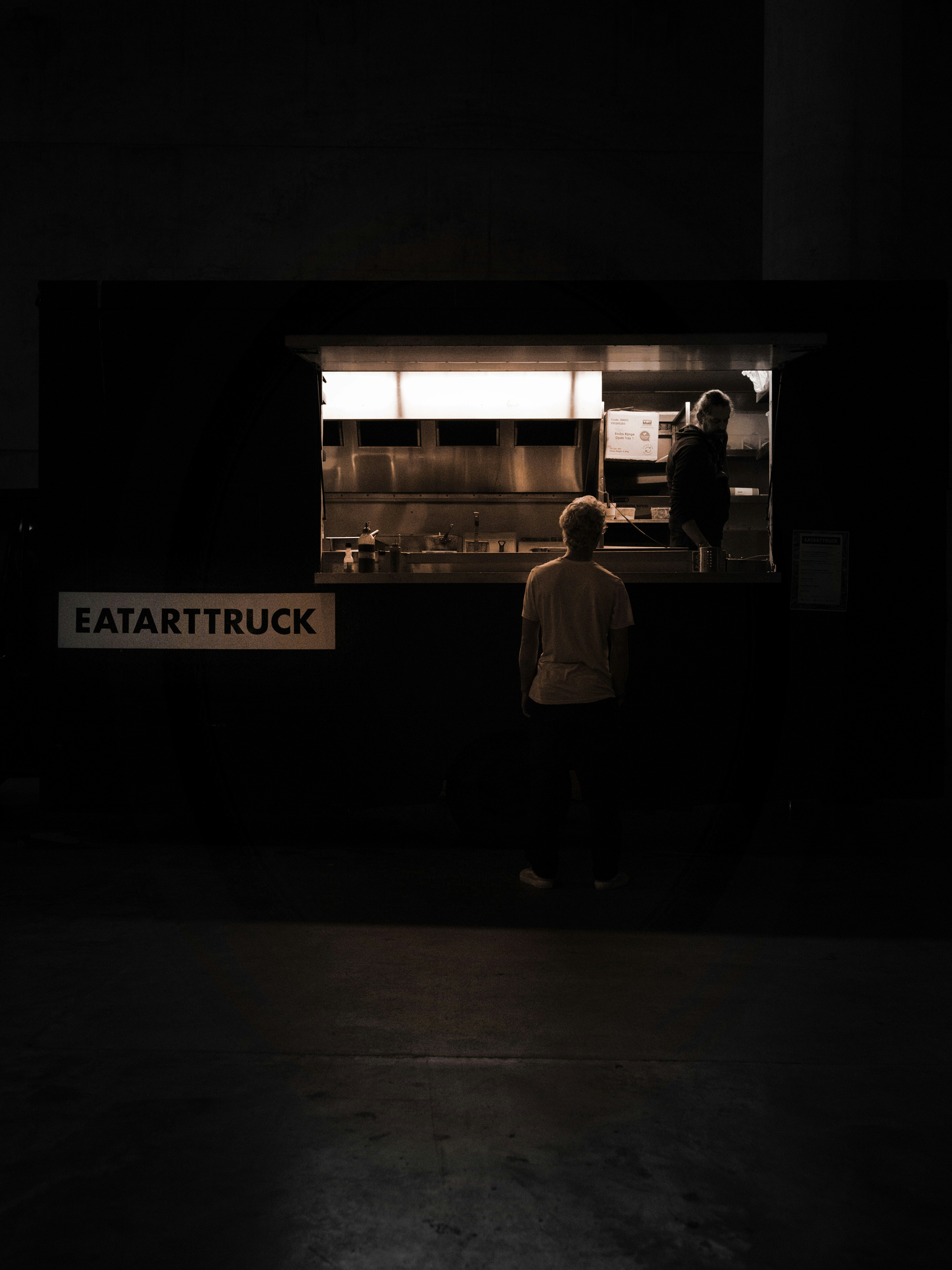 A food truck illuminated against a dark backdrop, with a customer waiting in anticipation. The scene captures the essence of late-night street dining.