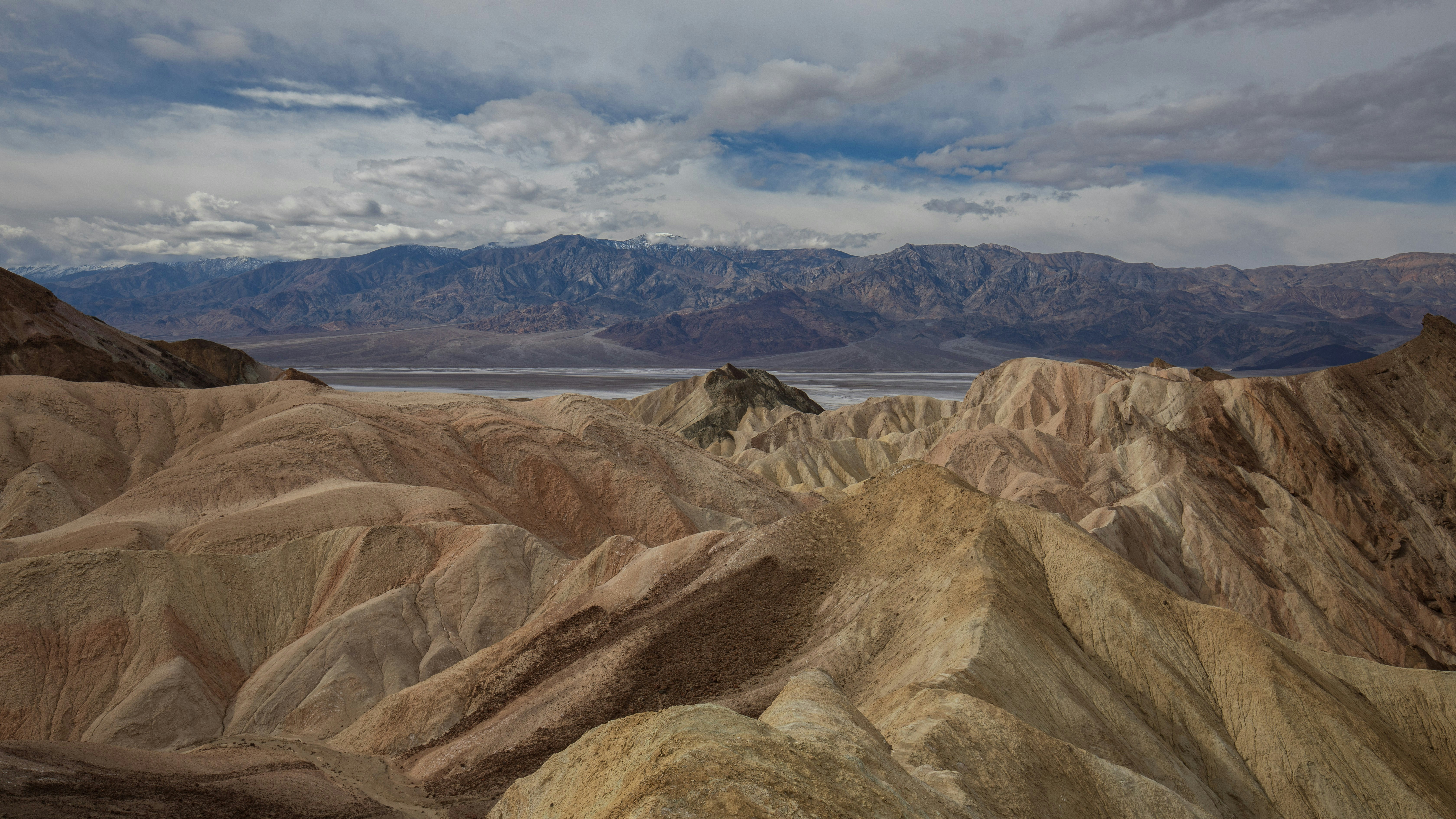 A sweeping view of layered, multicolored hills under a dramatic sky in a desert landscape. The image captures the intricate textures and patterns formed by natural erosion.