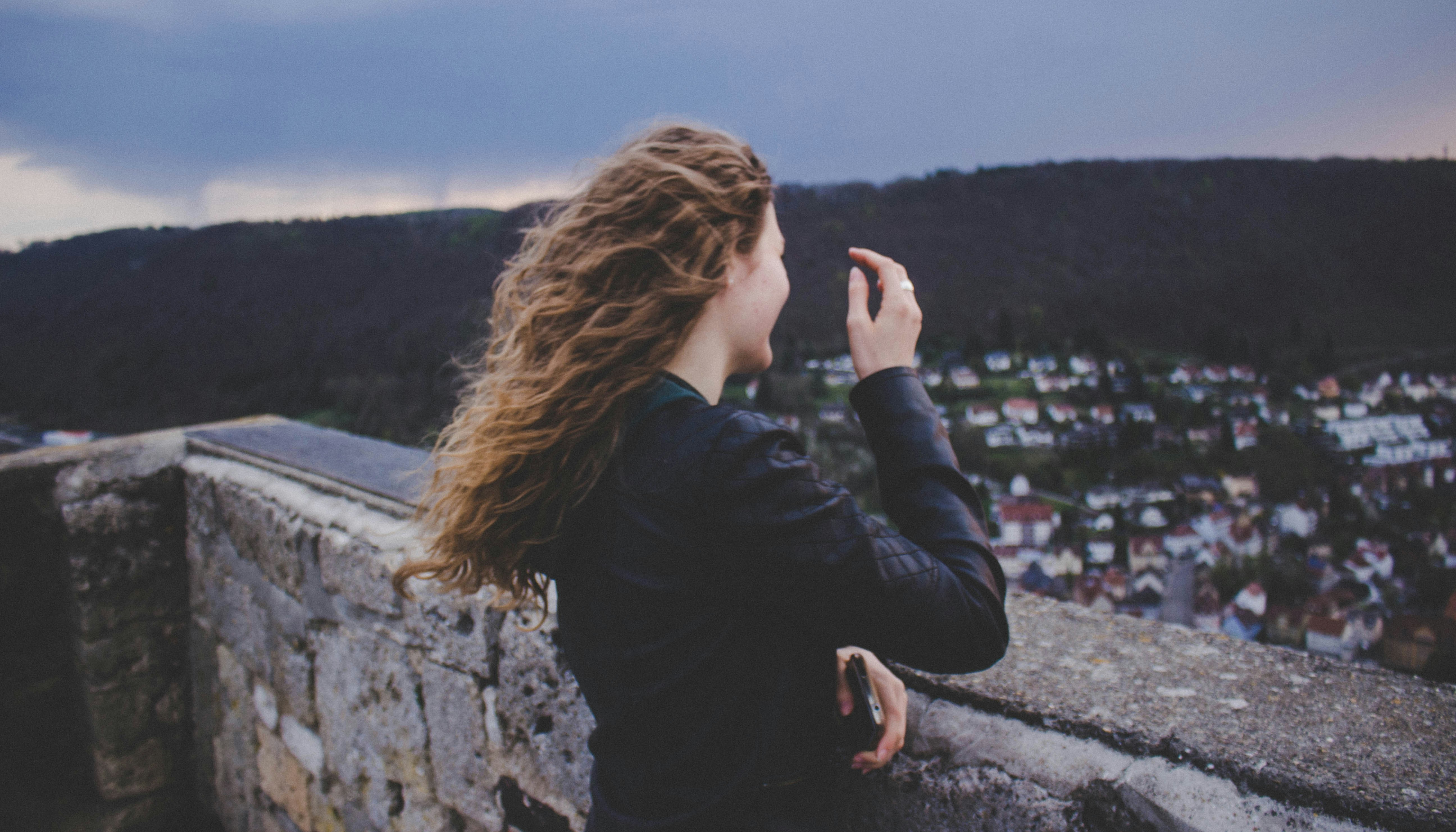 Woman with flowing hair gazes over a scenic landscape from a stone wall, capturing a moment of contemplation.