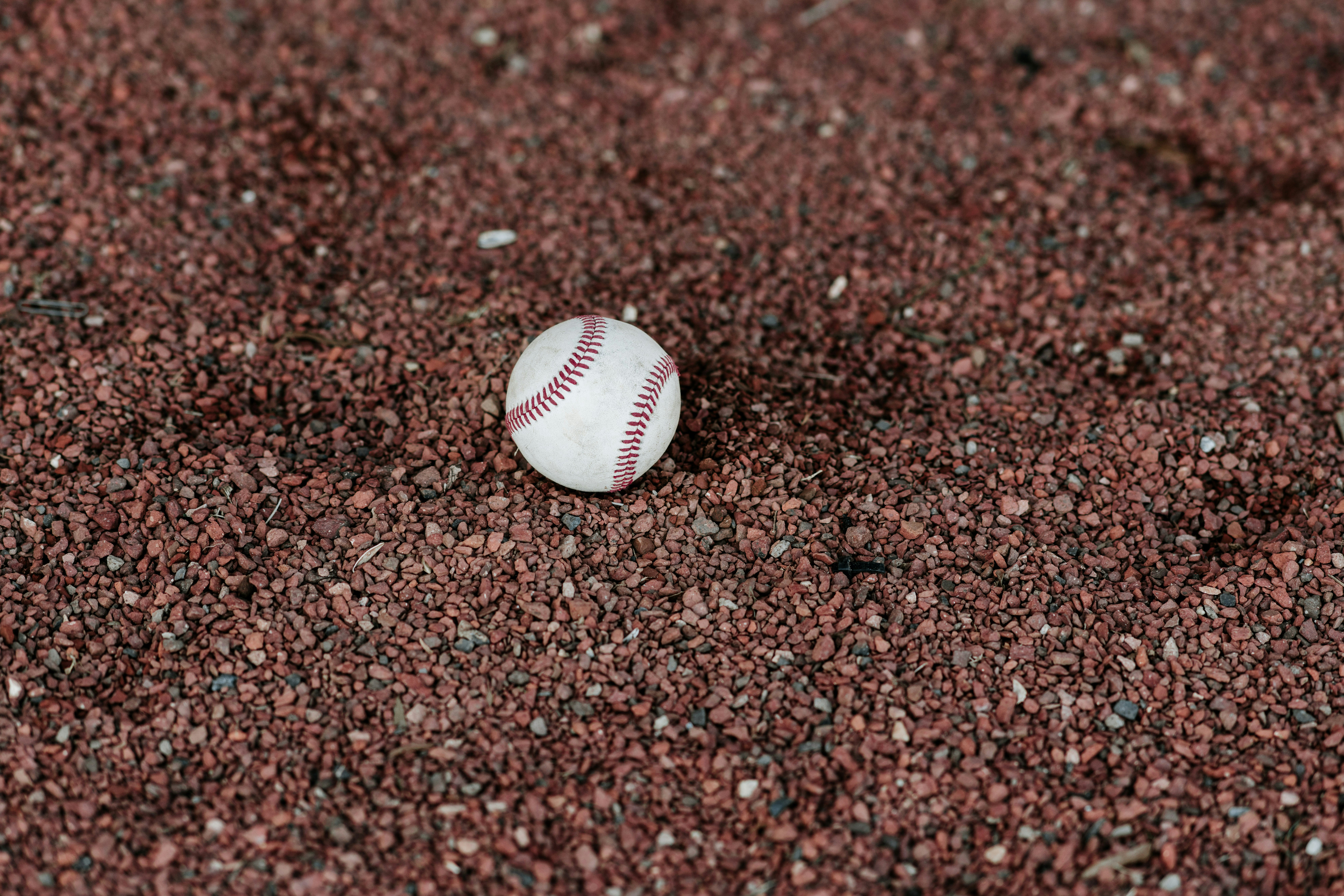 A single baseball rests on a bed of reddish gravel, highlighting its texture and the surrounding environment.