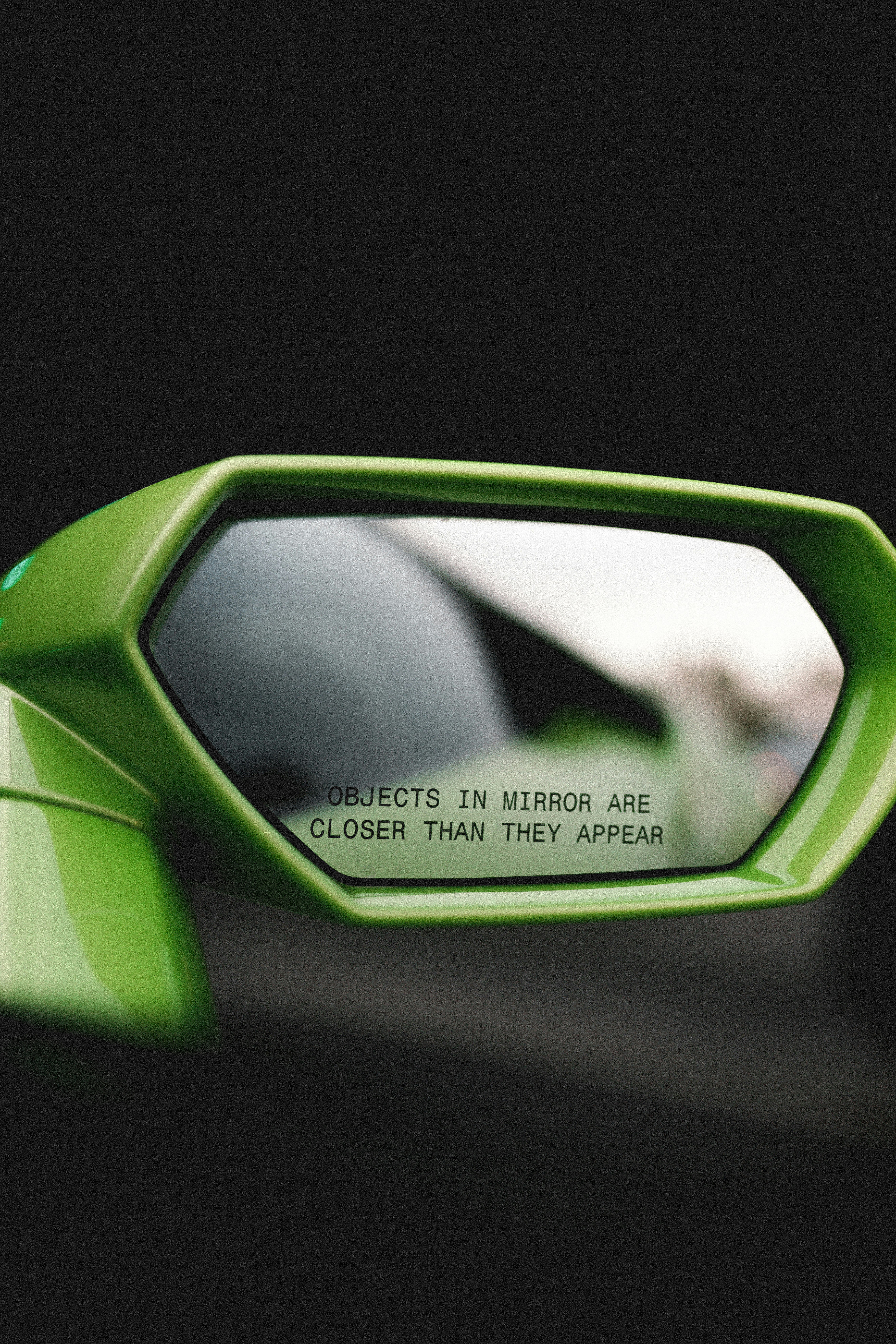 Side mirror displaying the warning 'OBJECTS IN MIRROR ARE CLOSER THAN THEY APPEAR' with a blurred background. The vibrant green frame adds a dynamic touch.