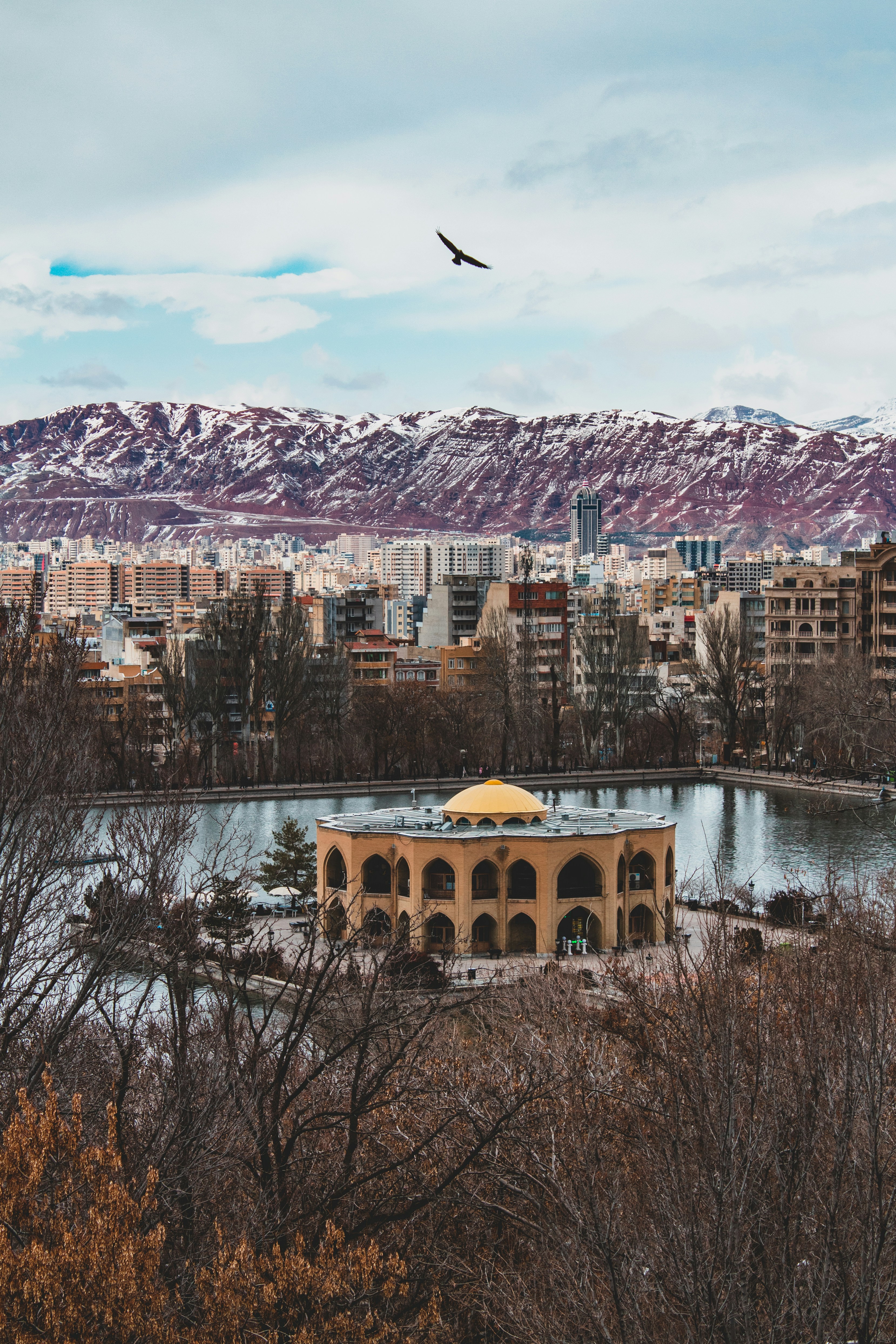 A historic pavilion surrounded by a serene lake, framed by mountains and urban architecture. A bird soars above, adding to the tranquil atmosphere.