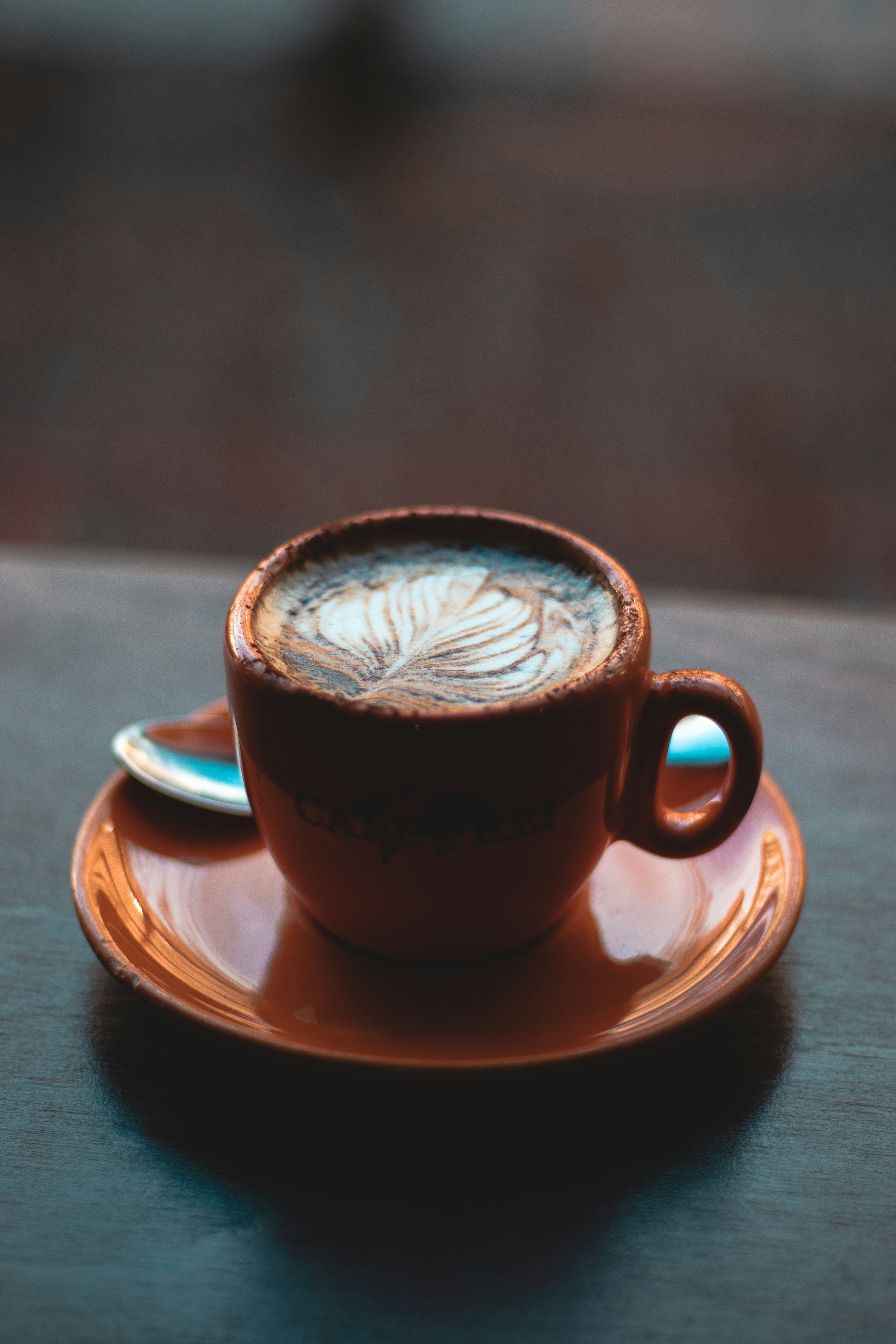 A beautifully crafted latte with intricate foam art sits on a vibrant orange saucer, accompanied by a small spoon. The warm tones evoke a cozy café atmosphere.