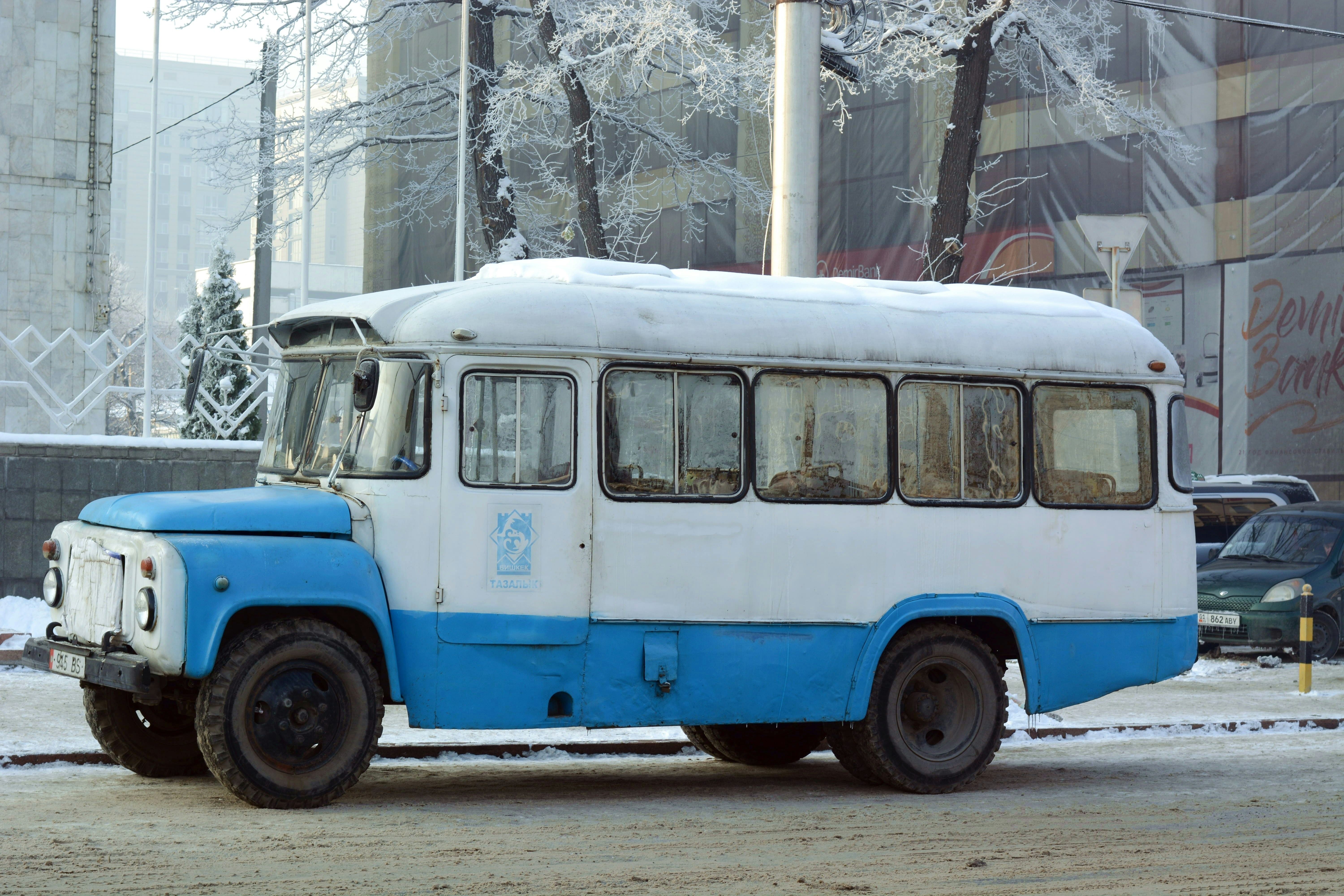 A vintage blue and white bus covered in snow, parked on a winter street surrounded by frosty trees and buildings.