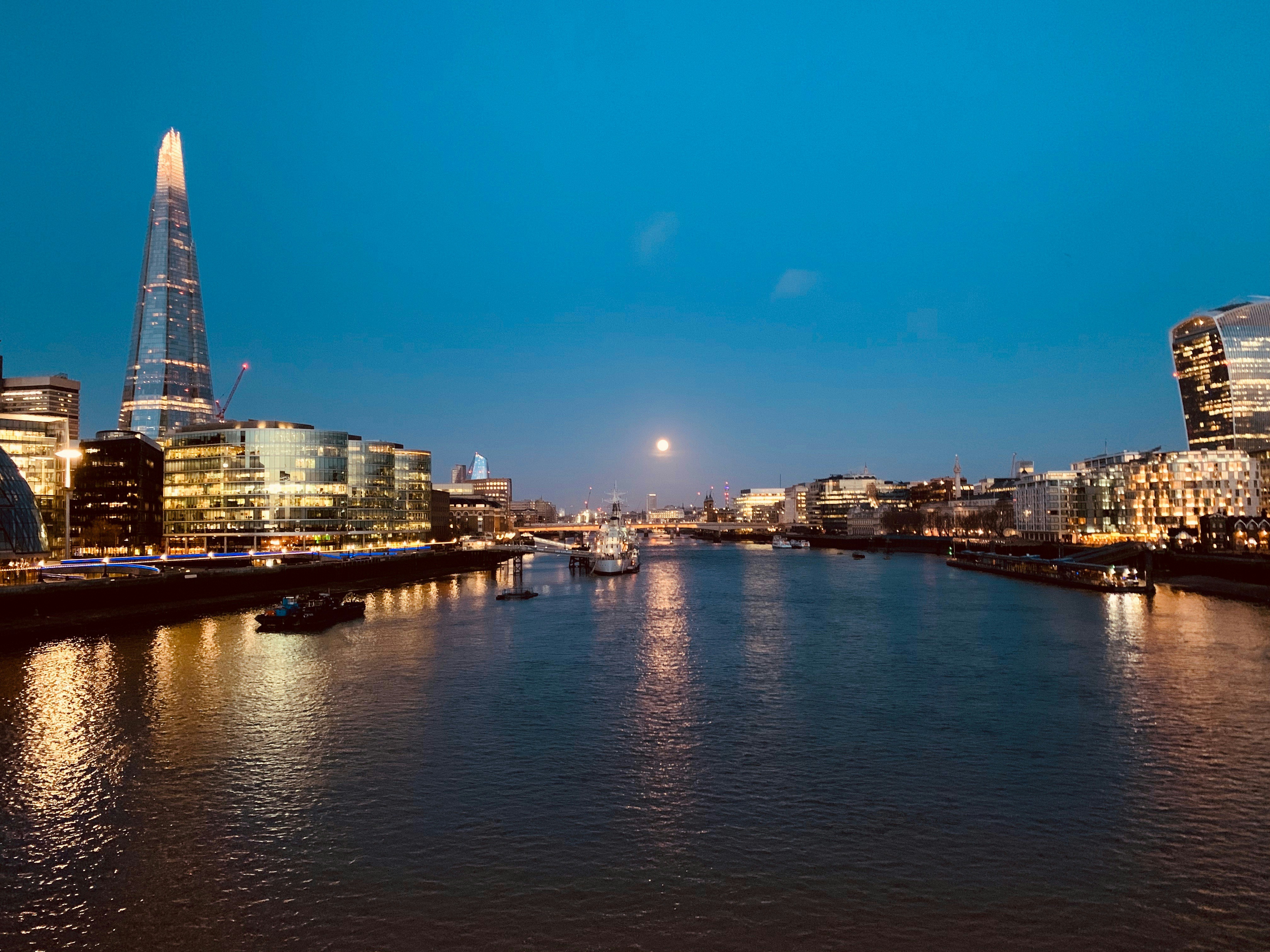 The Shard towers over the Thames River, illuminated by city lights, with a full moon casting reflections on the water's surface.