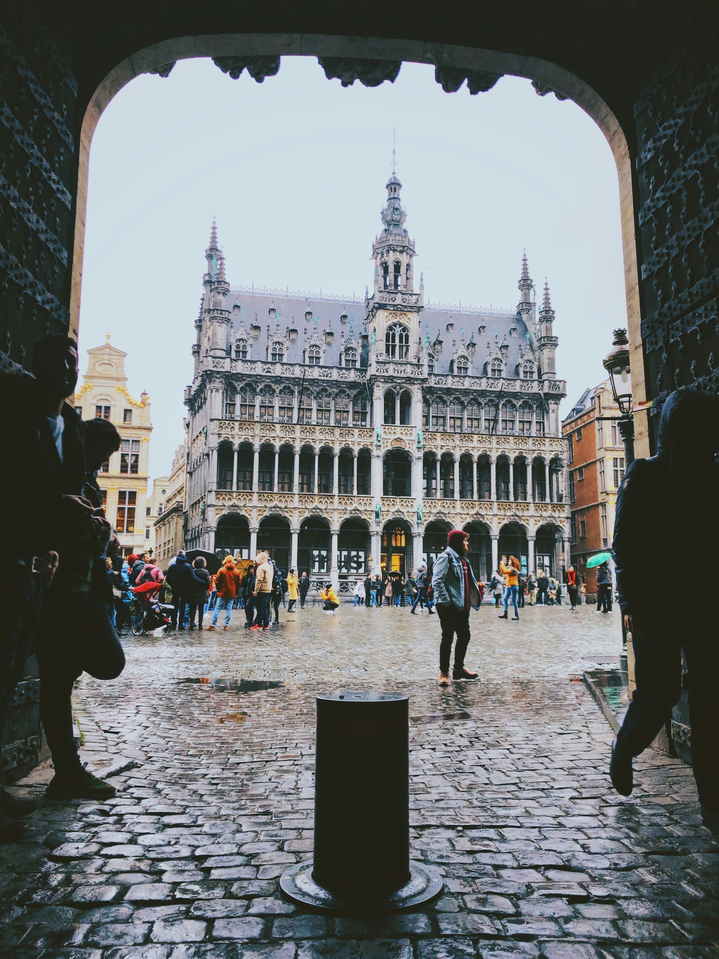 Historic building in Brussels framed by an archway, bustling with visitors under a cloudy sky. A central column stands in the foreground.
