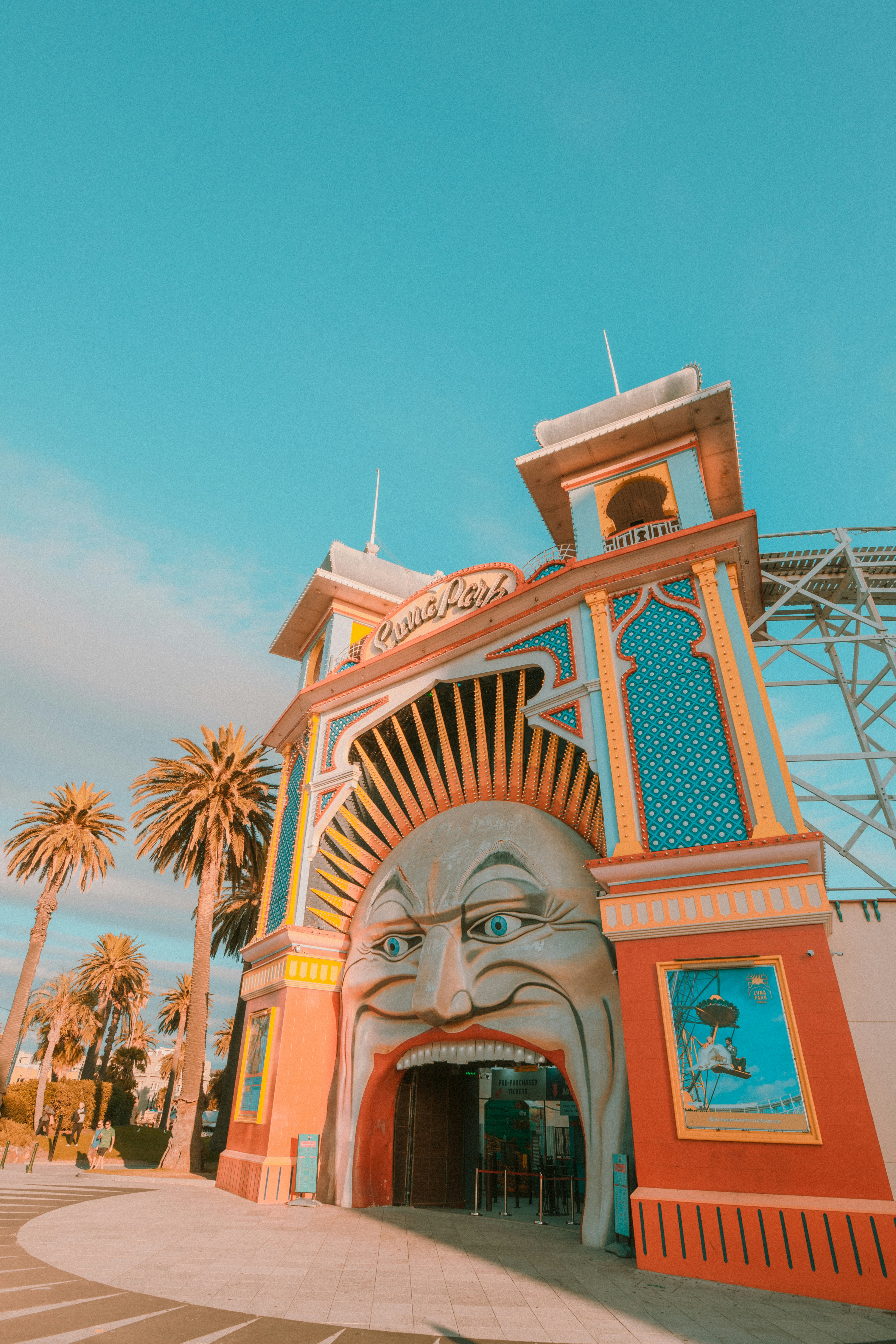 Vibrant funhouse entrance featuring a large, expressive face and colorful decorations, framed by palm trees under a clear blue sky.
