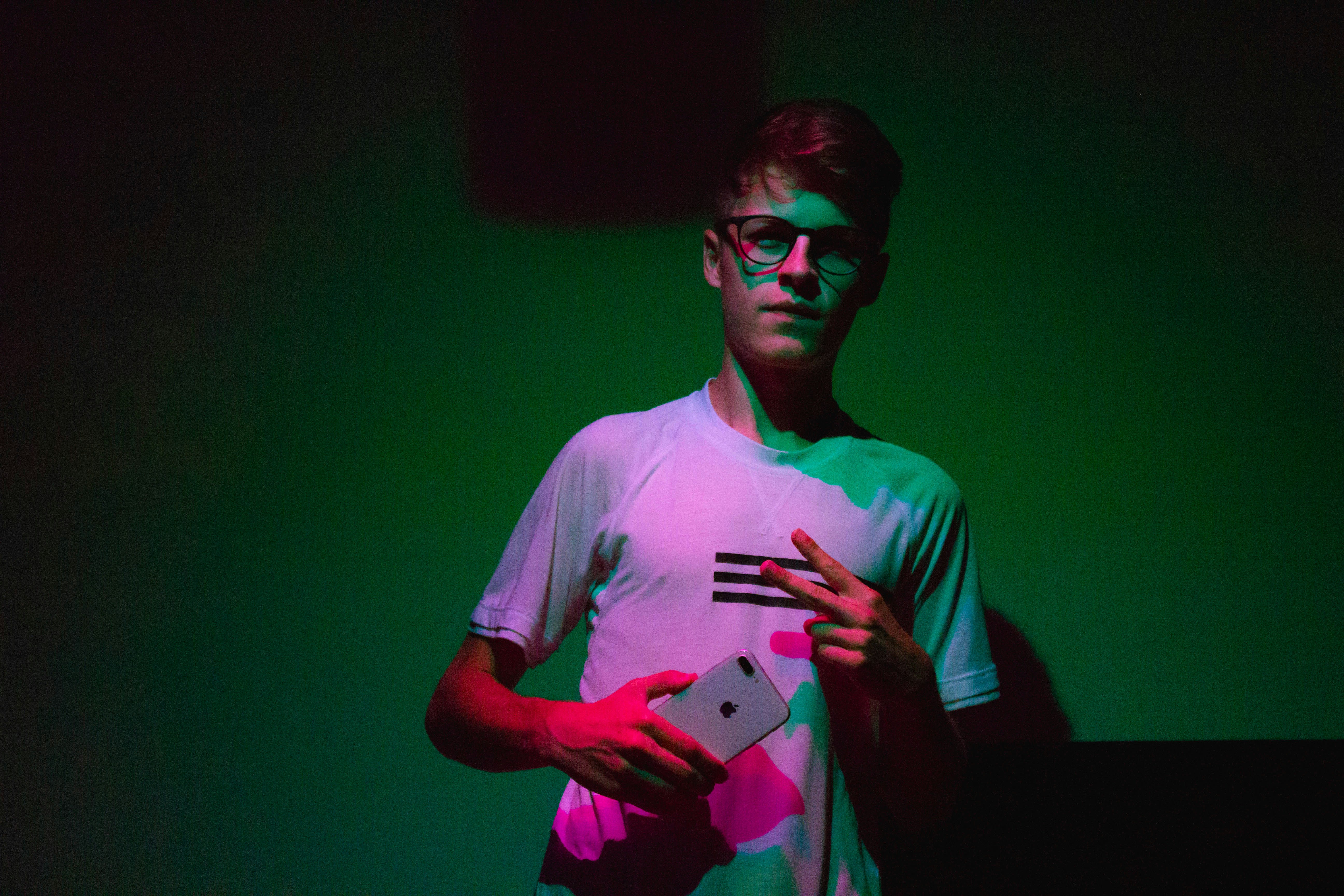 Young man with glasses posing against a green backdrop, holding a smartphone and making a peace sign.