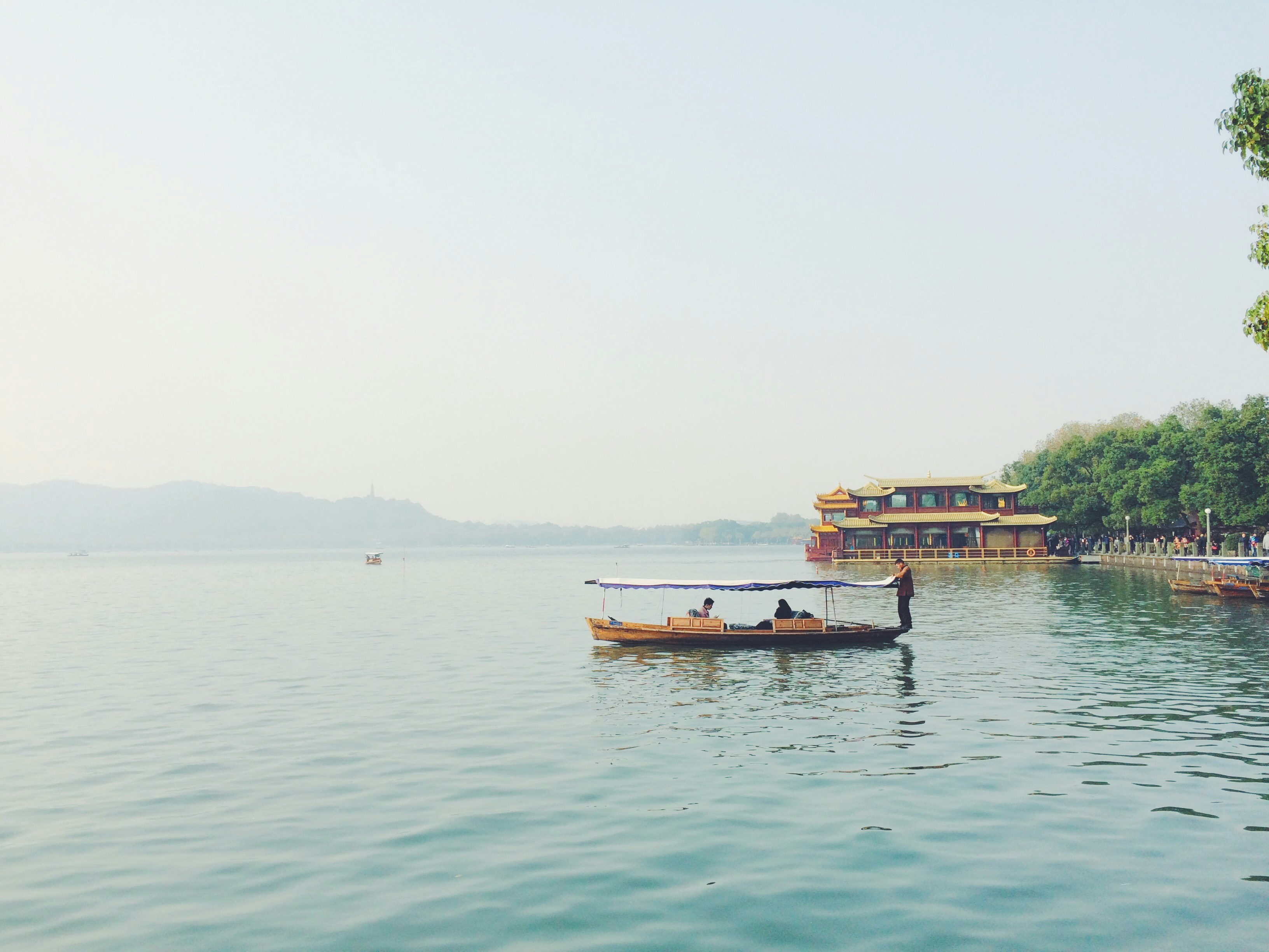 Small boat gliding on a calm lake with misty mountains and a distant pavilion.