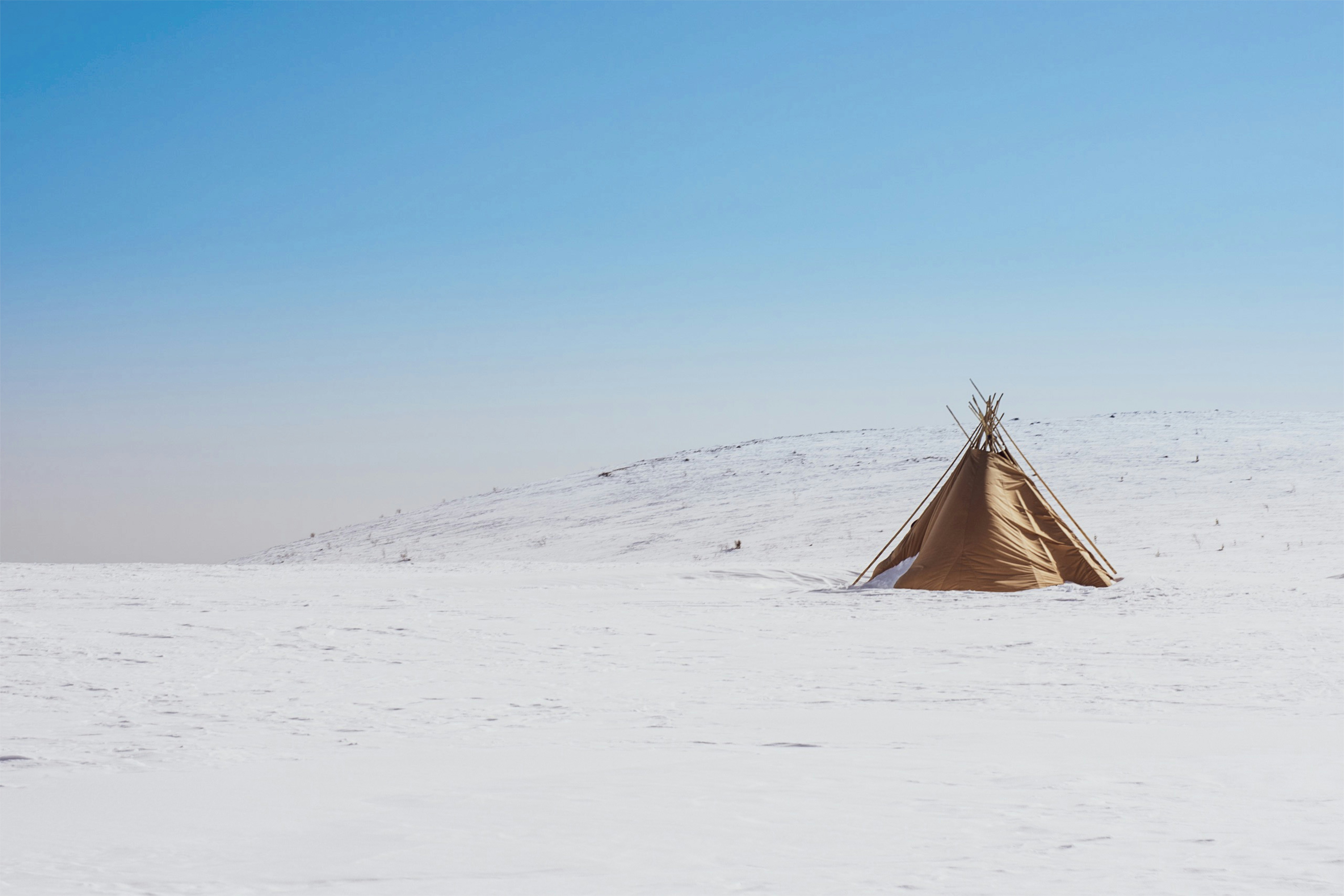 A traditional teepee stands alone on a vast, snowy landscape under a clear blue sky.