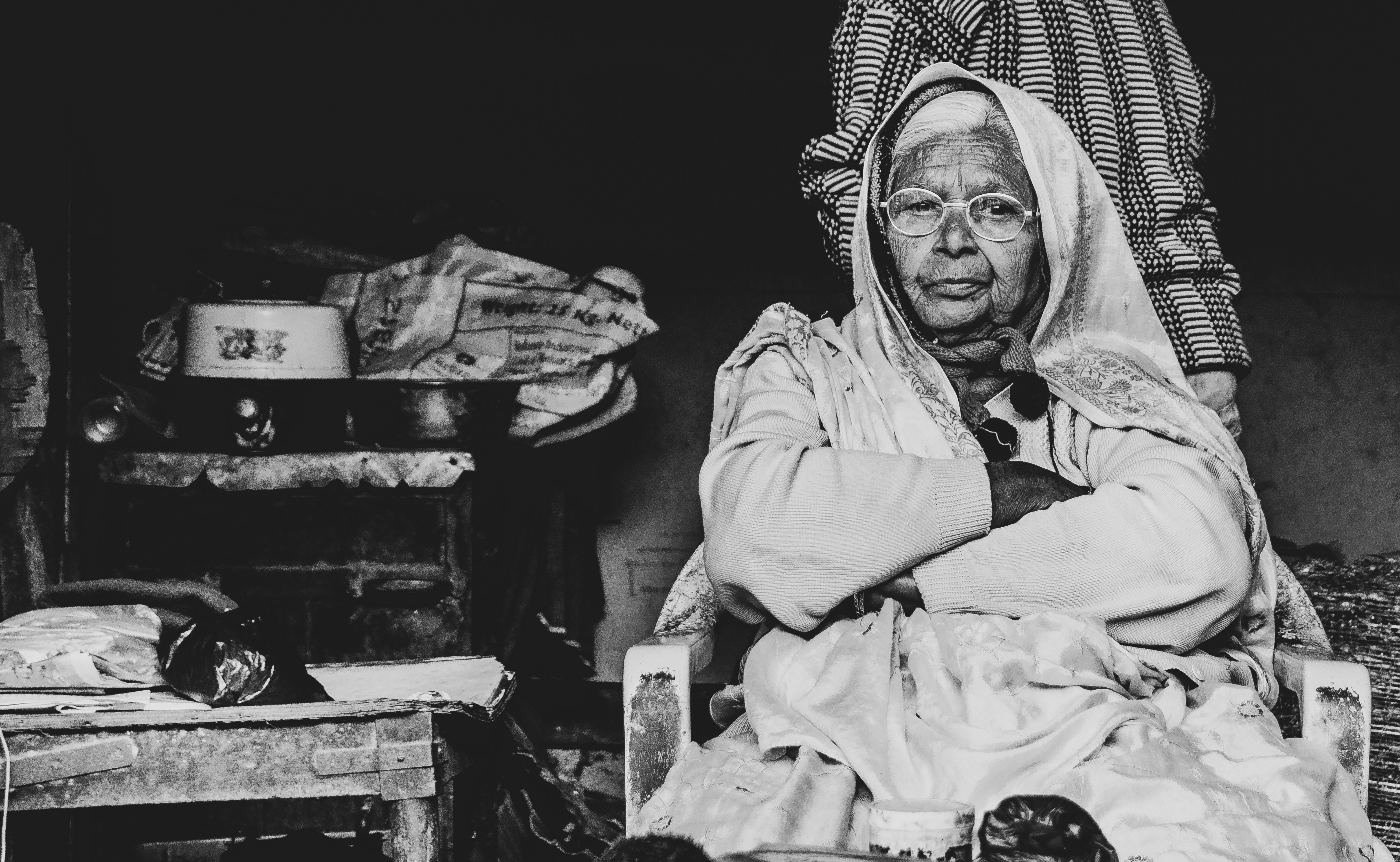 Elderly person sitting in a dimly lit room with various belongings around, captured in black and white.
