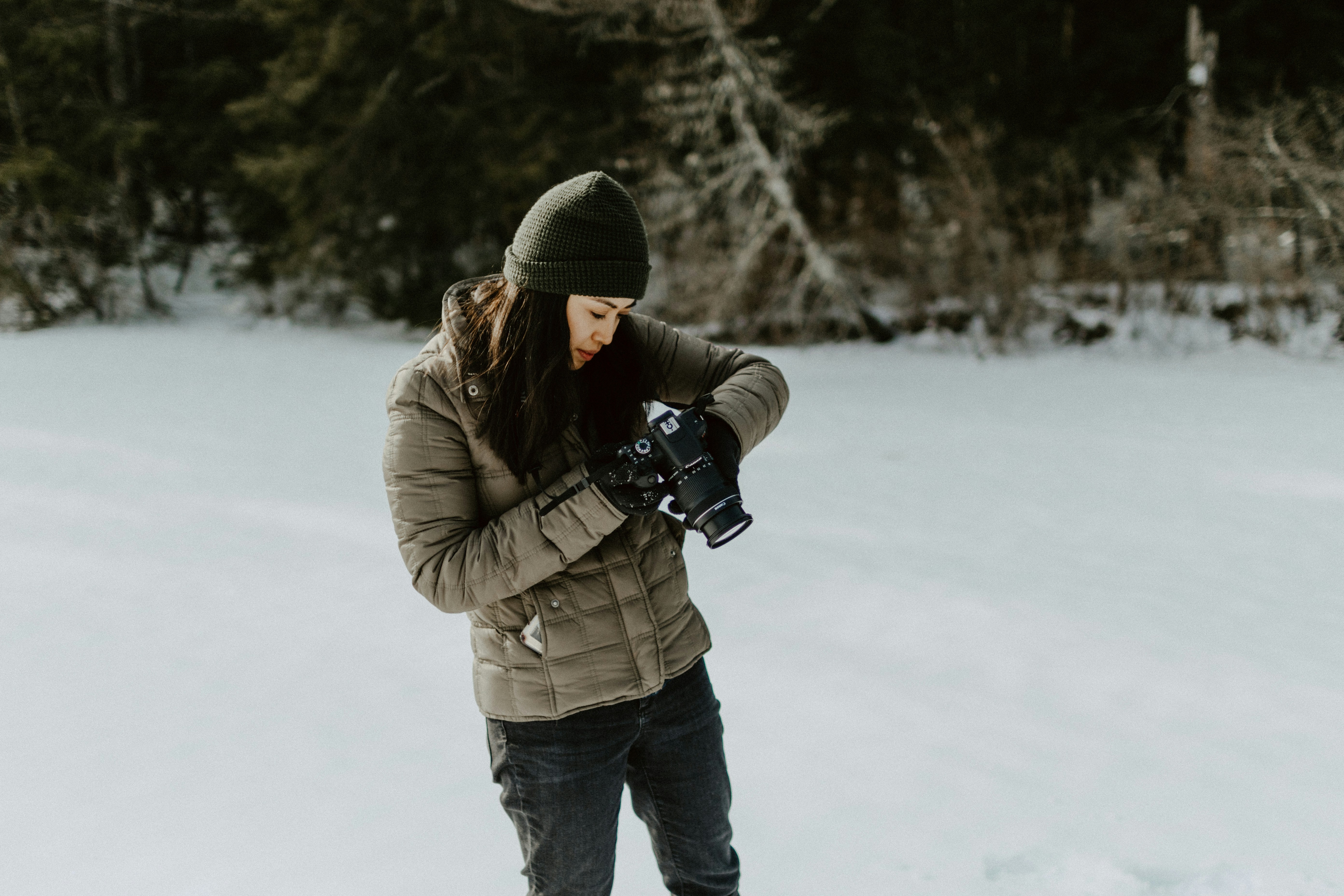 A photographer in winter attire checks her camera settings while standing on a snowy landscape, surrounded by trees.