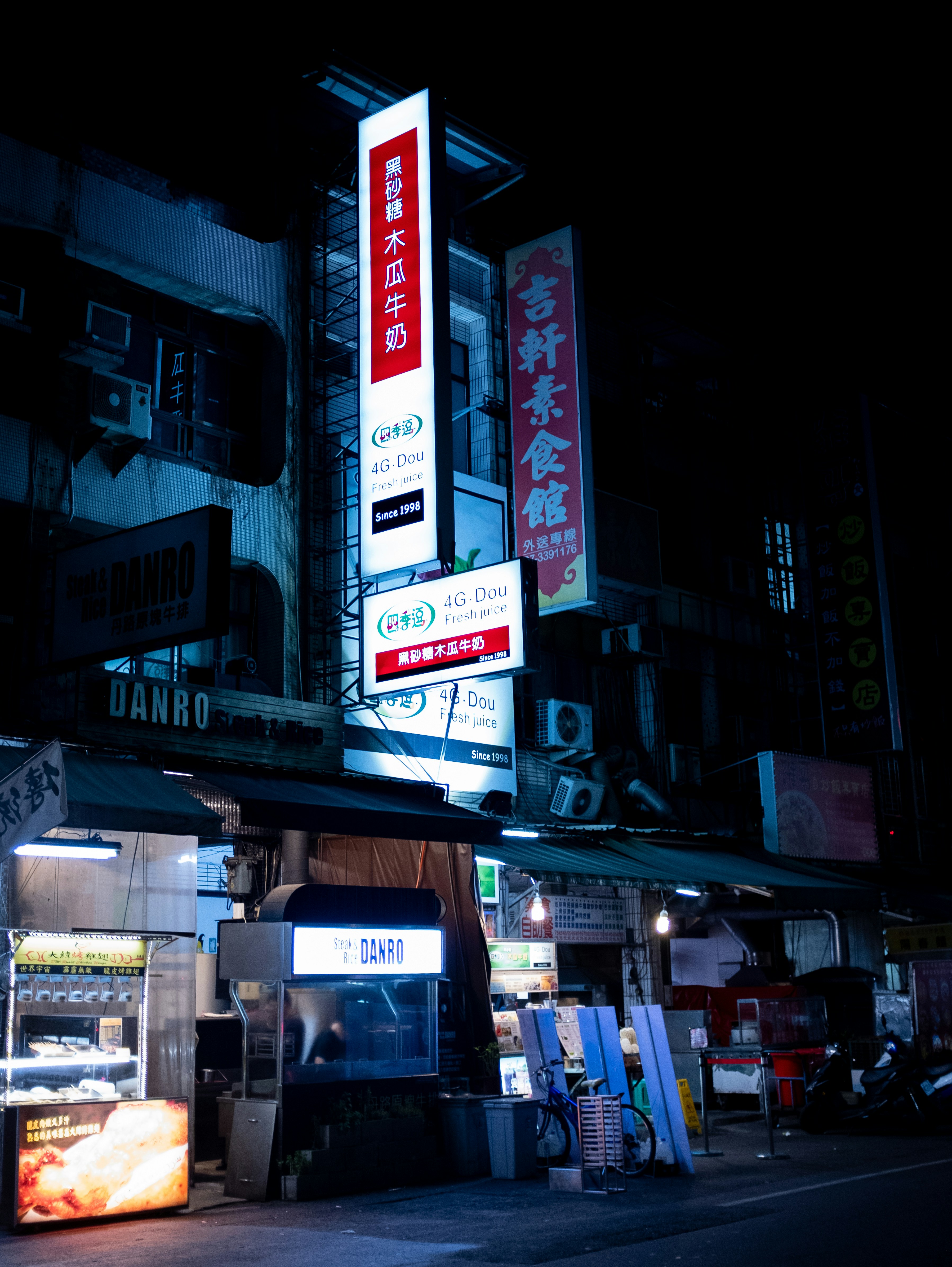 Illuminated storefronts of a night market, showcasing vibrant signage and a bustling atmosphere under the evening sky.