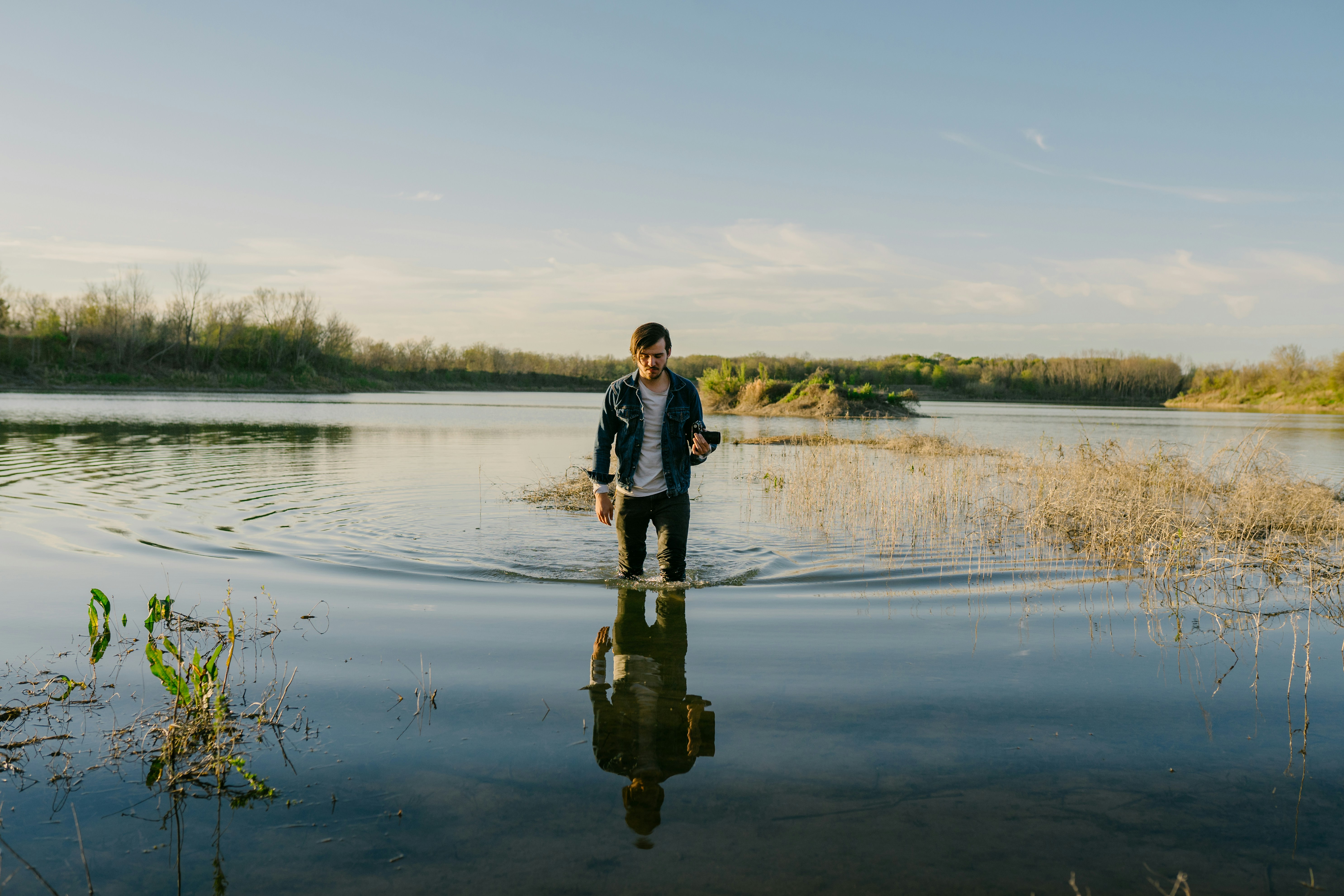 Person wading through shallow water, surrounded by a serene landscape and gentle ripples. Their reflection is visible in the calm surface.