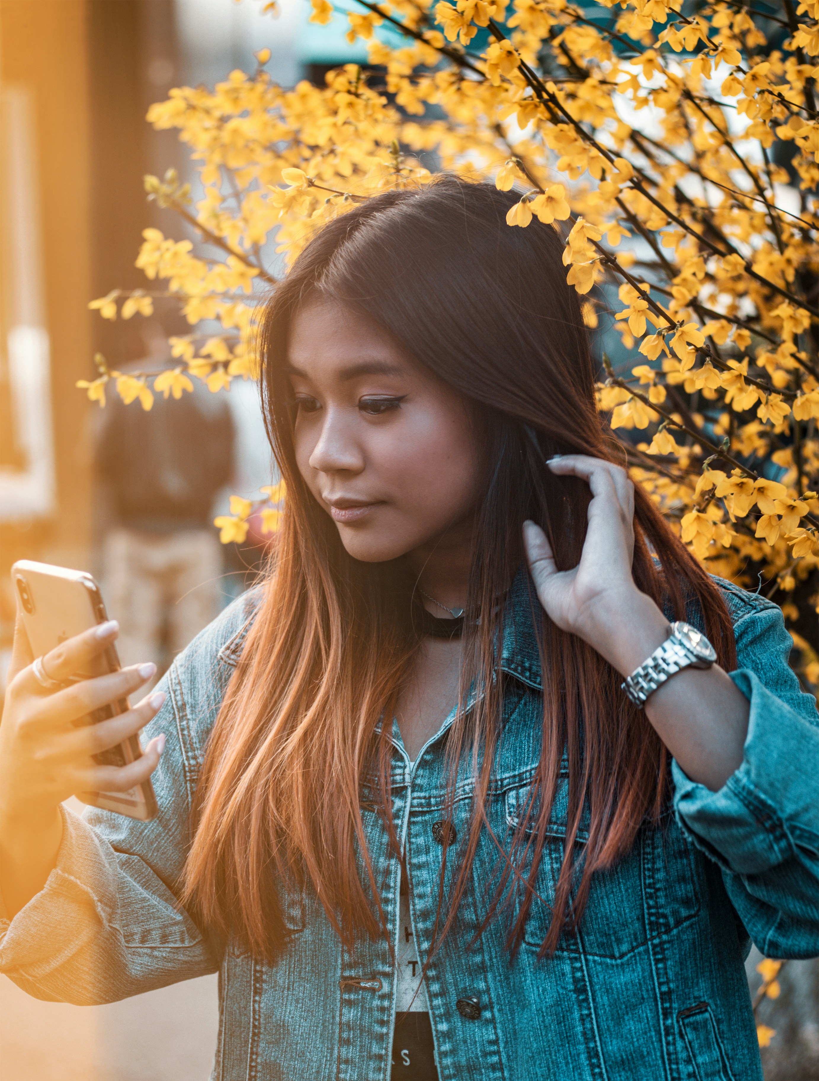 Young woman in a denim jacket interacts with her smartphone, surrounded by vibrant yellow flowers. The scene captures a blend of technology and nature.