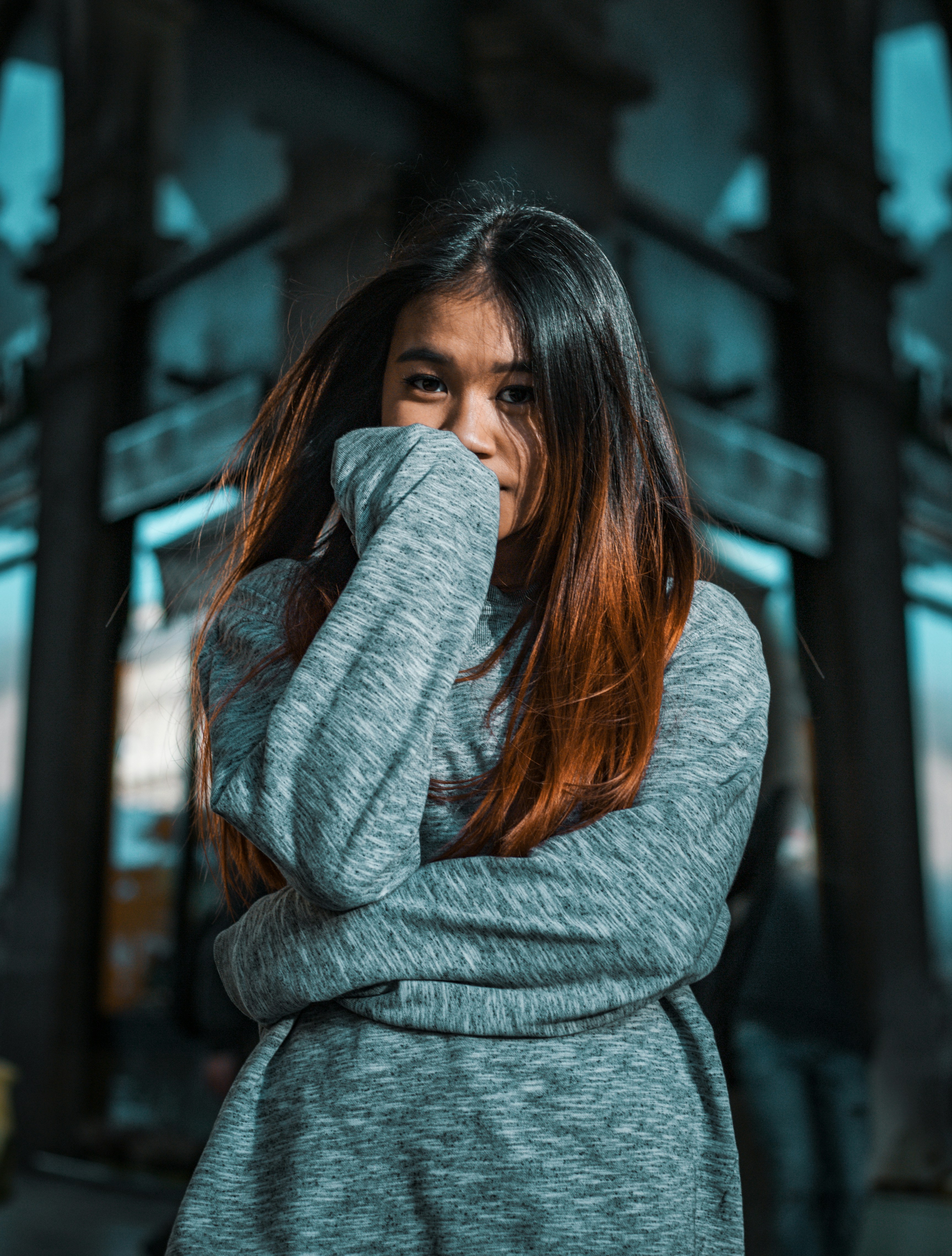 A young woman stands in a city setting, partially concealing her face with her sweater, evoking a sense of introspection amidst the bustling backdrop.