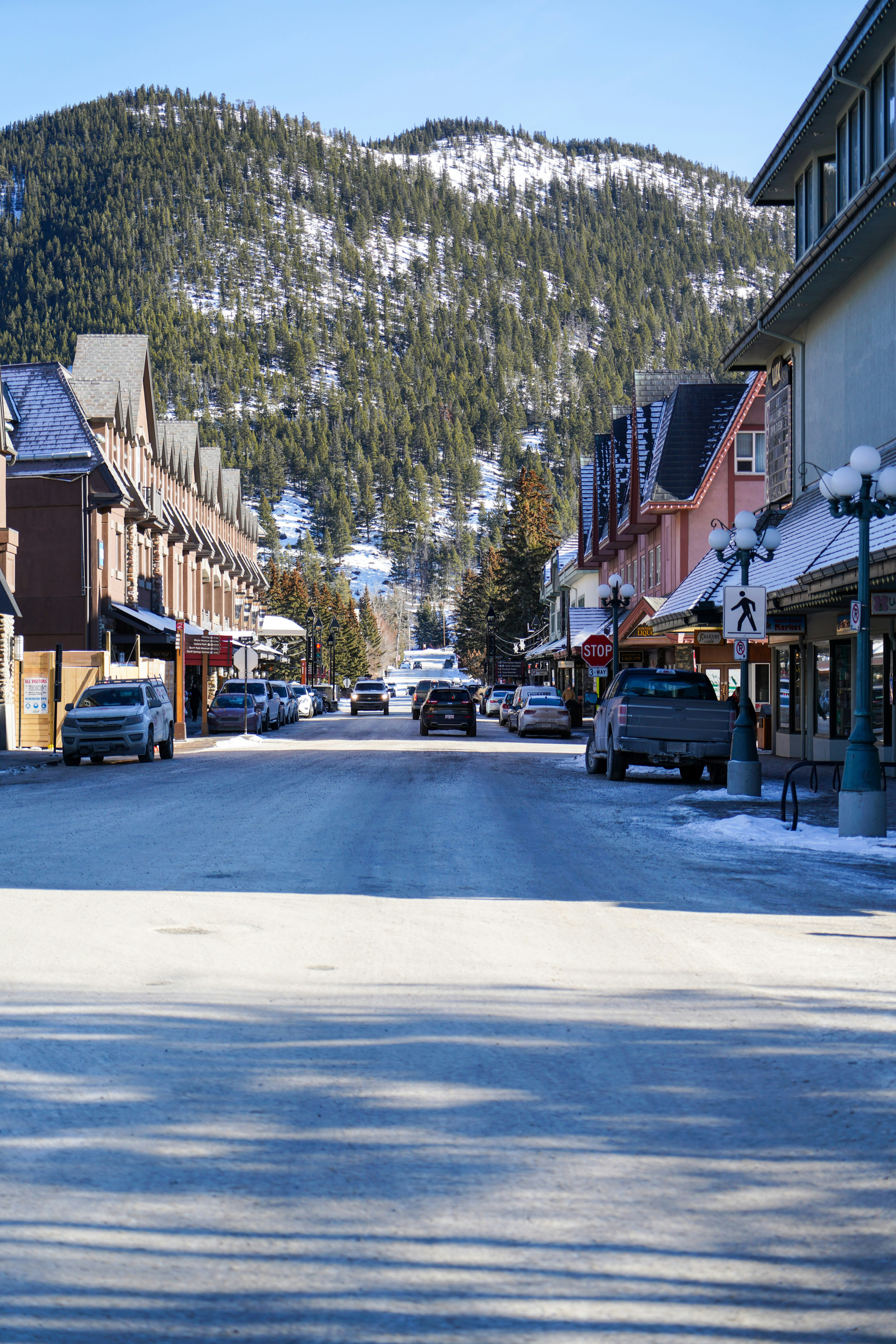 Snow-covered street lined with quaint shops and vehicles, framed by majestic mountains in the background.