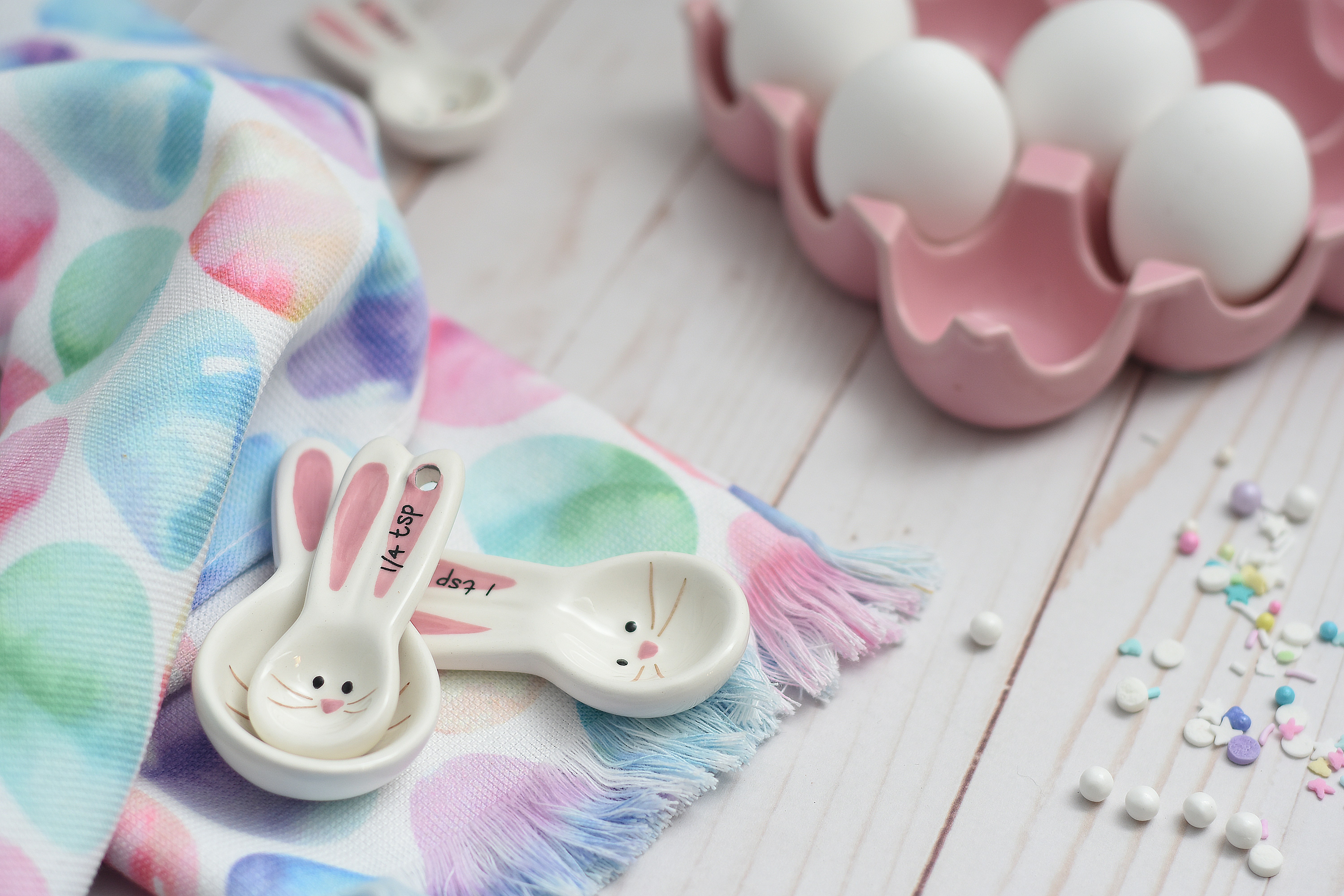 Ceramic bunny measuring spoons on a colorful pastel cloth with white eggs in a pink holder.