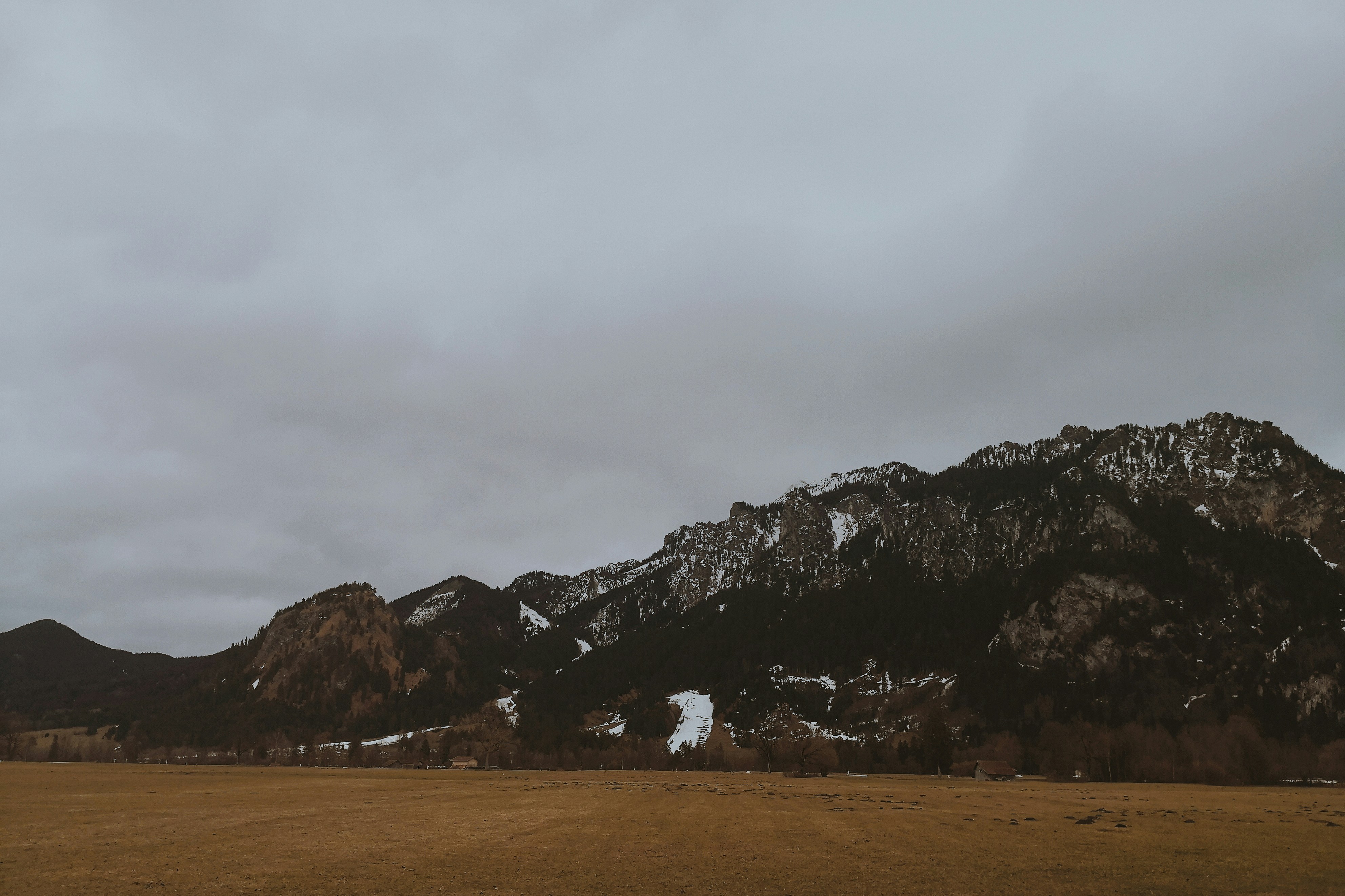 Snow-dusted granite peaks rise above a flat, golden-brown field under a heavy gray sky. The composition emphasizes the rugged silhouette against an expansive plain.